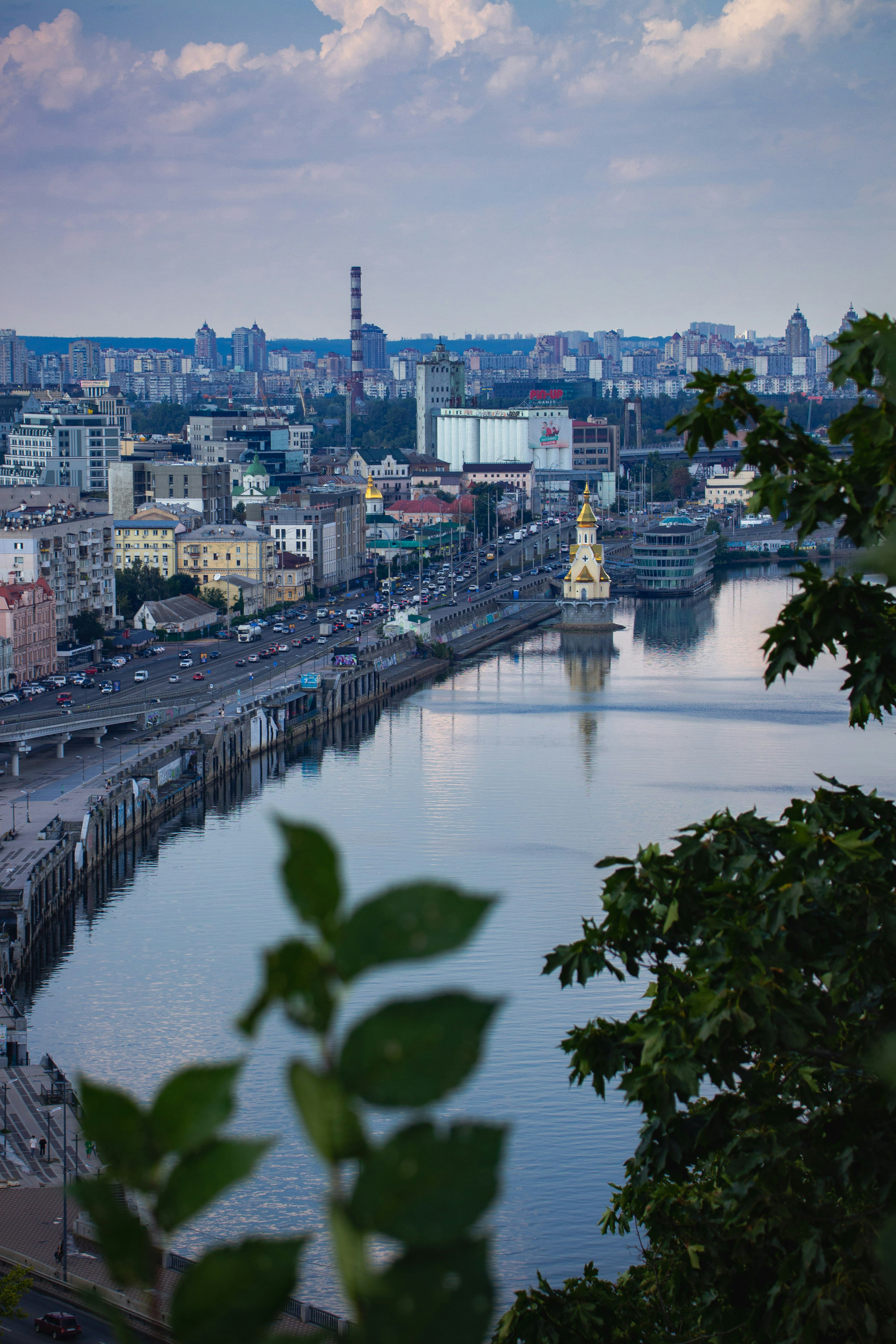 A panoramic view of a bustling cityscape along a river, showcasing a blend of modern architecture and natural elements. The scene is framed by greenery in the foreground.