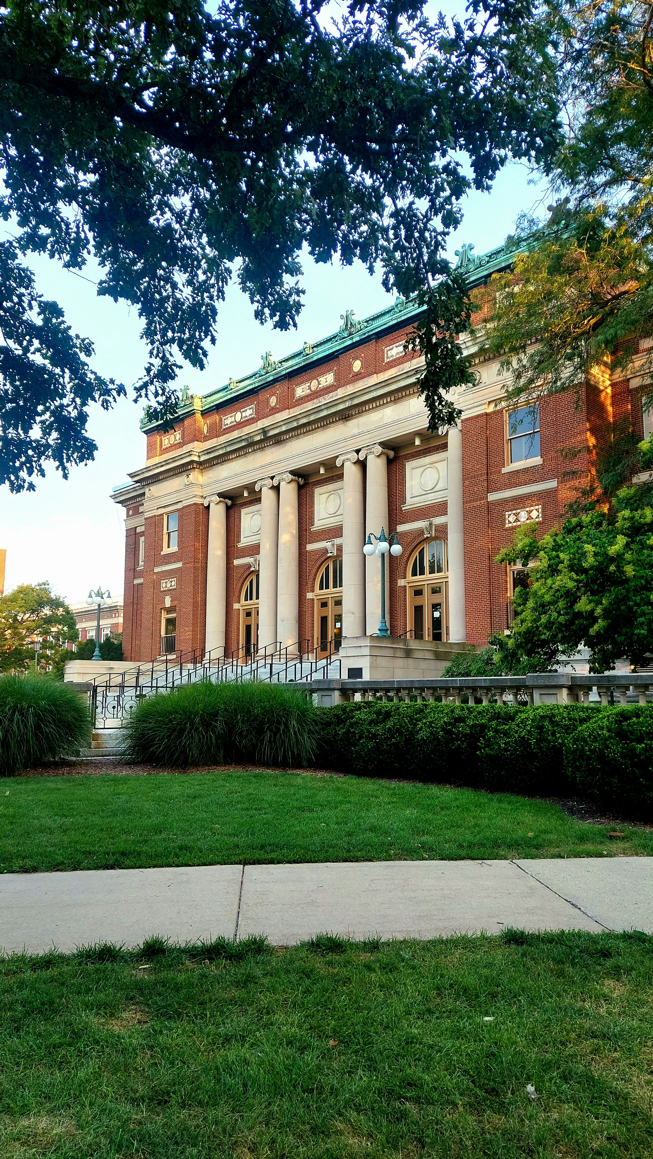 Elegant red-brick building with classical columns surrounded by lush greenery and trees under a blue sky.