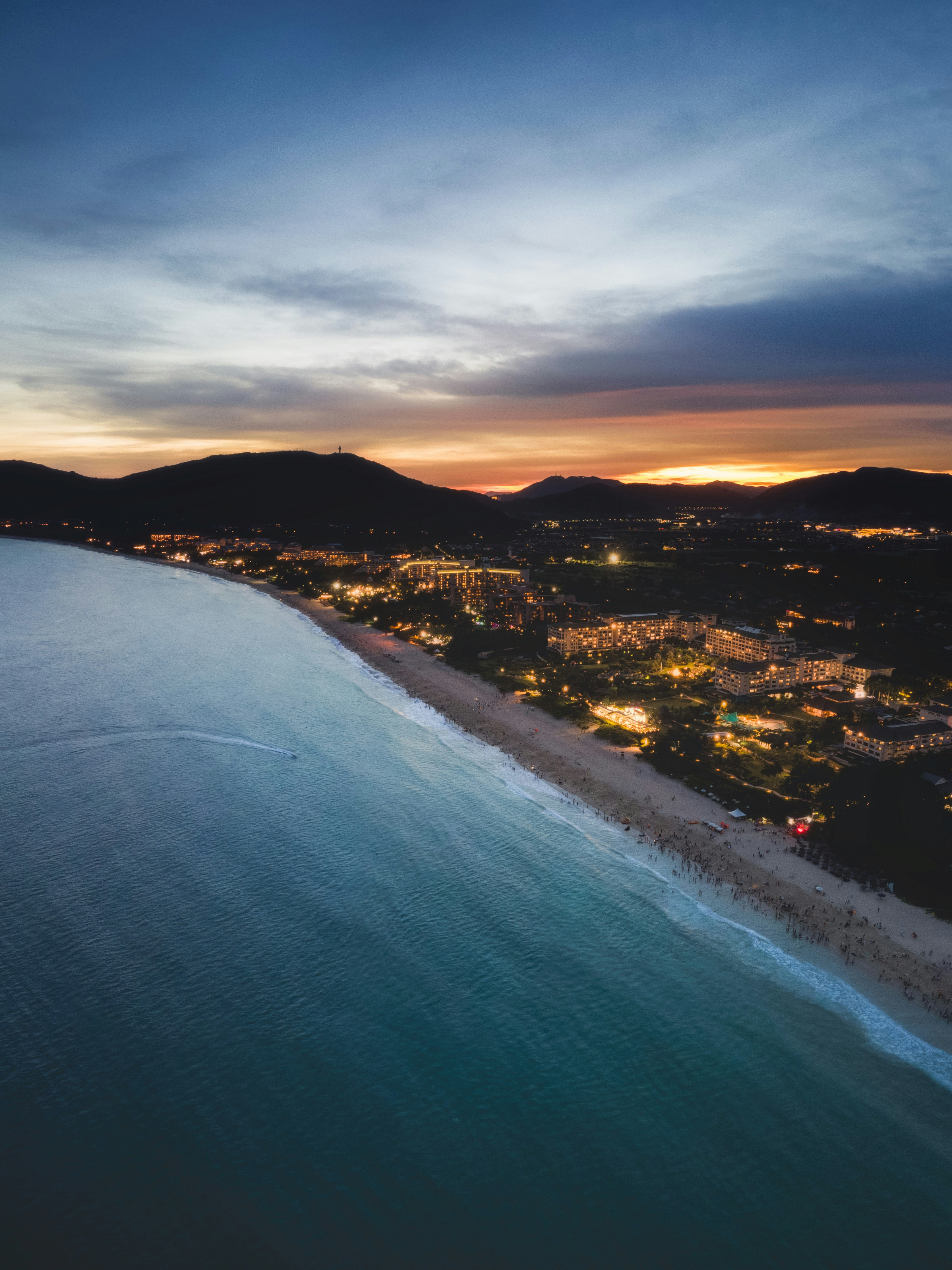 Aerial view of a beach at twilight, with glimmering lights from nearby resorts and a tranquil sea reflecting the dusk sky.