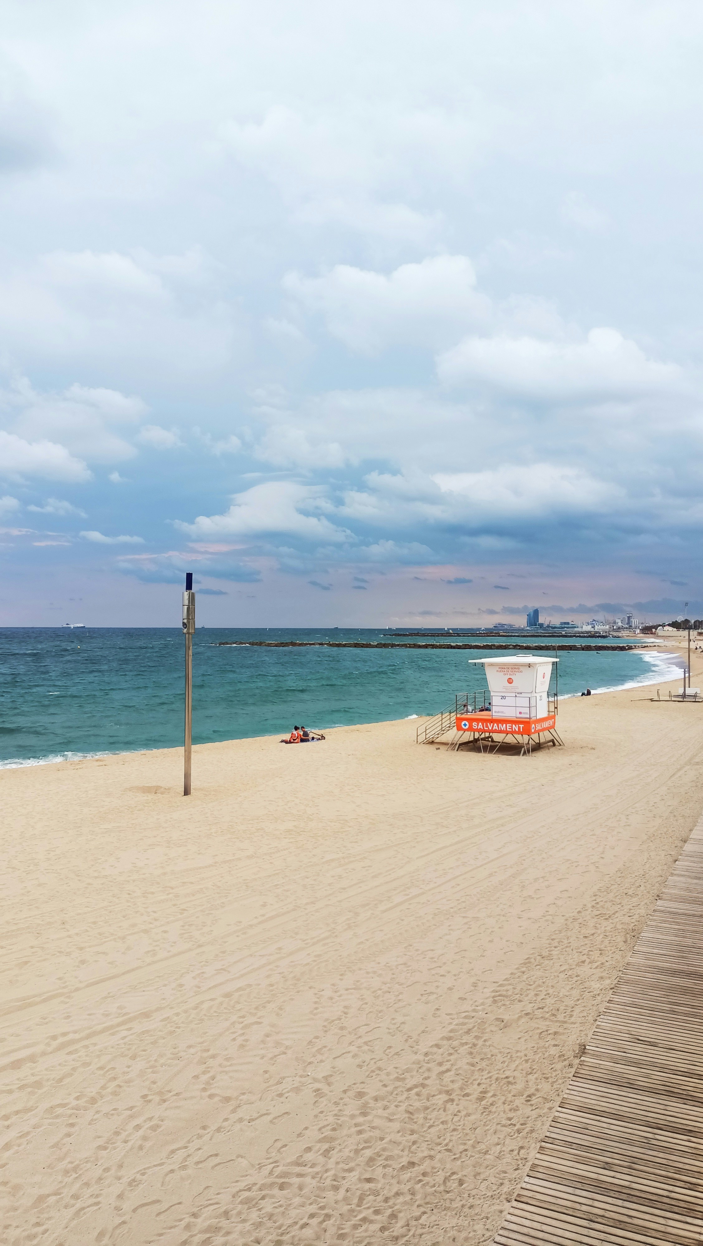 Photograph of a sandy beach with a lifeguard hut near the water, a wooden boardwalk on the right, and a dramatic cloud-filled sky.