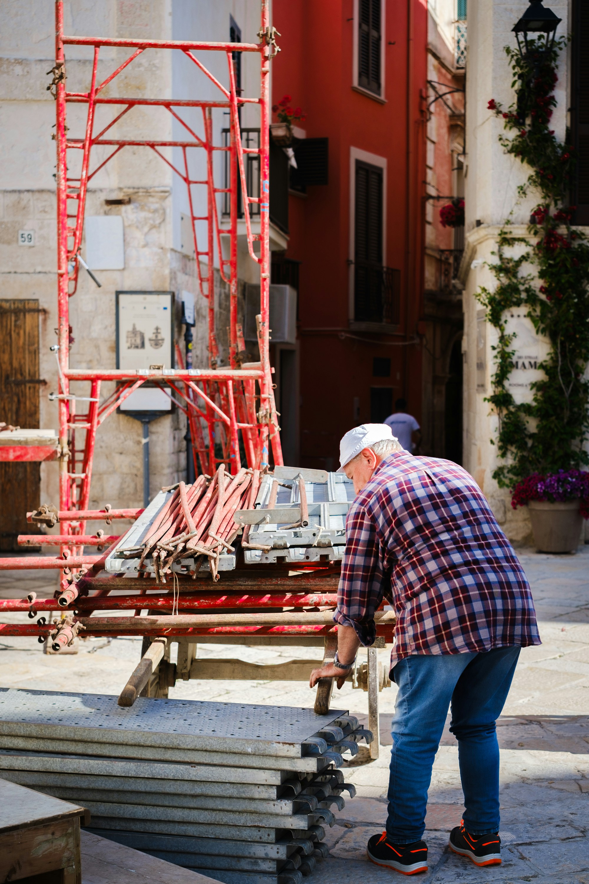 Worker in Plaid Shirt Rolls Scaffolding Amid Narrow Mediterranean Street