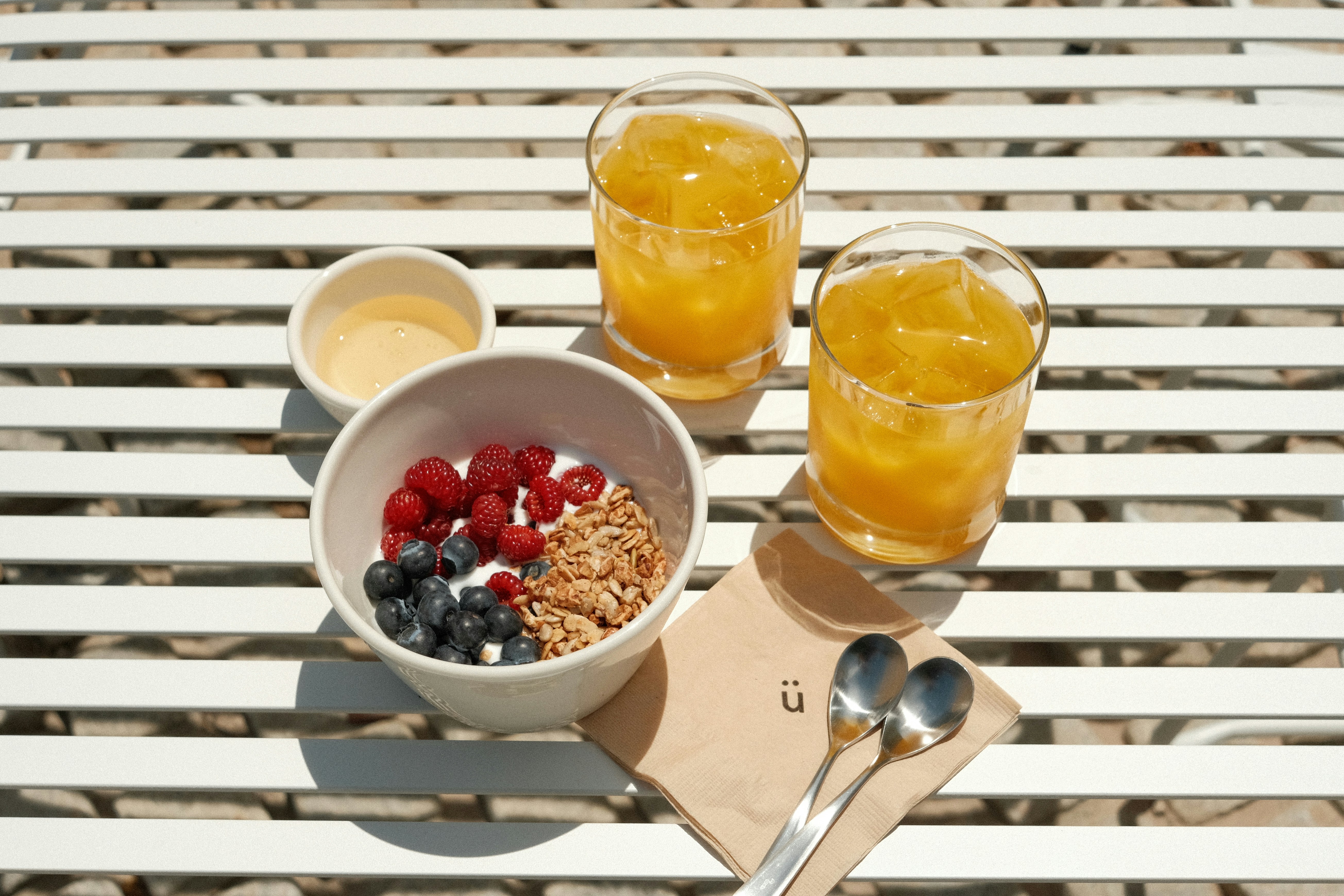 Sunlit Brunch Spread: Berry Granola Bowl with Two Glasses of Orange Juice on Slatted Table