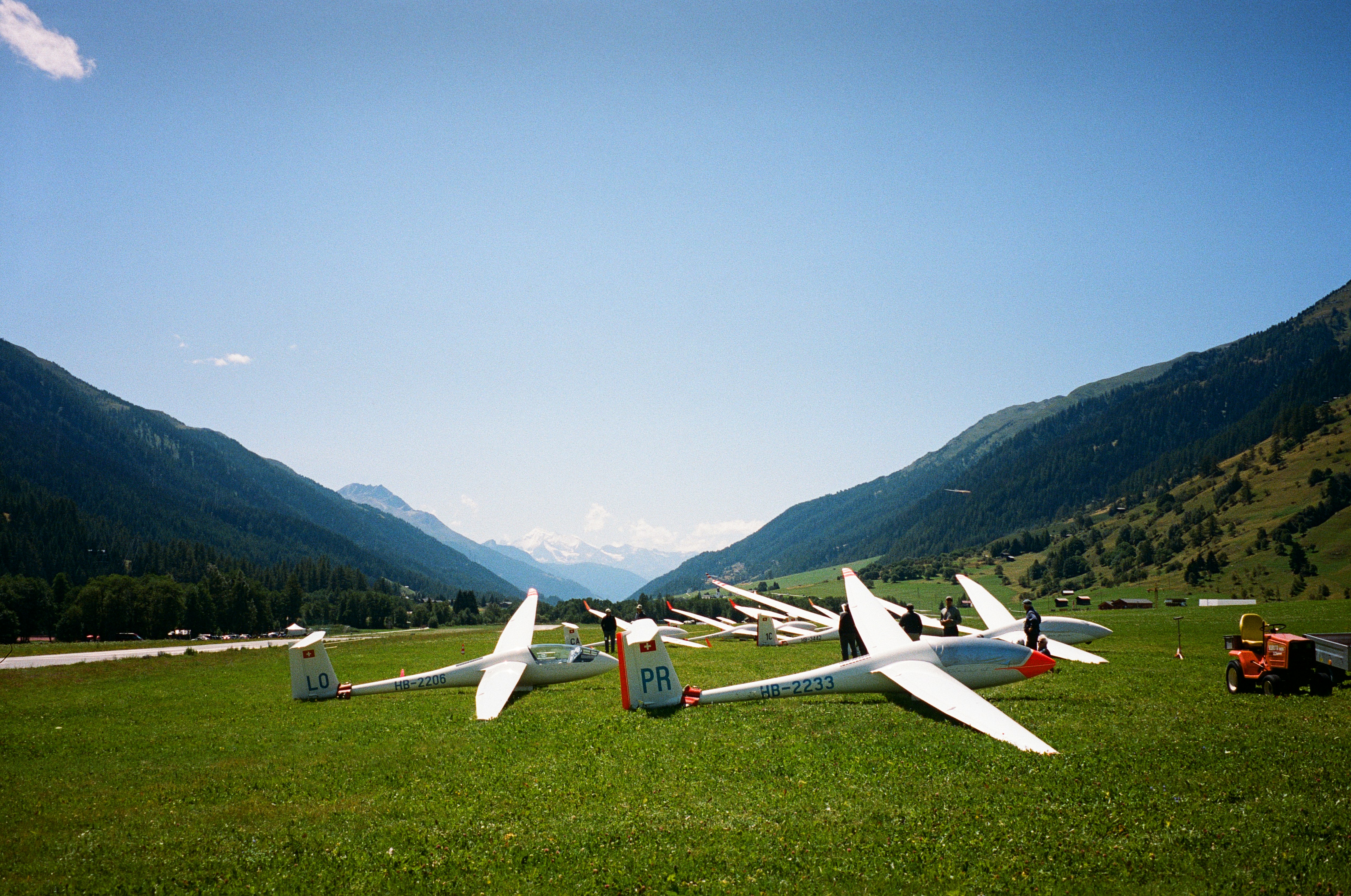 Gliders rest on a sunlit alpine meadow with distant mountains and a clear blue sky.