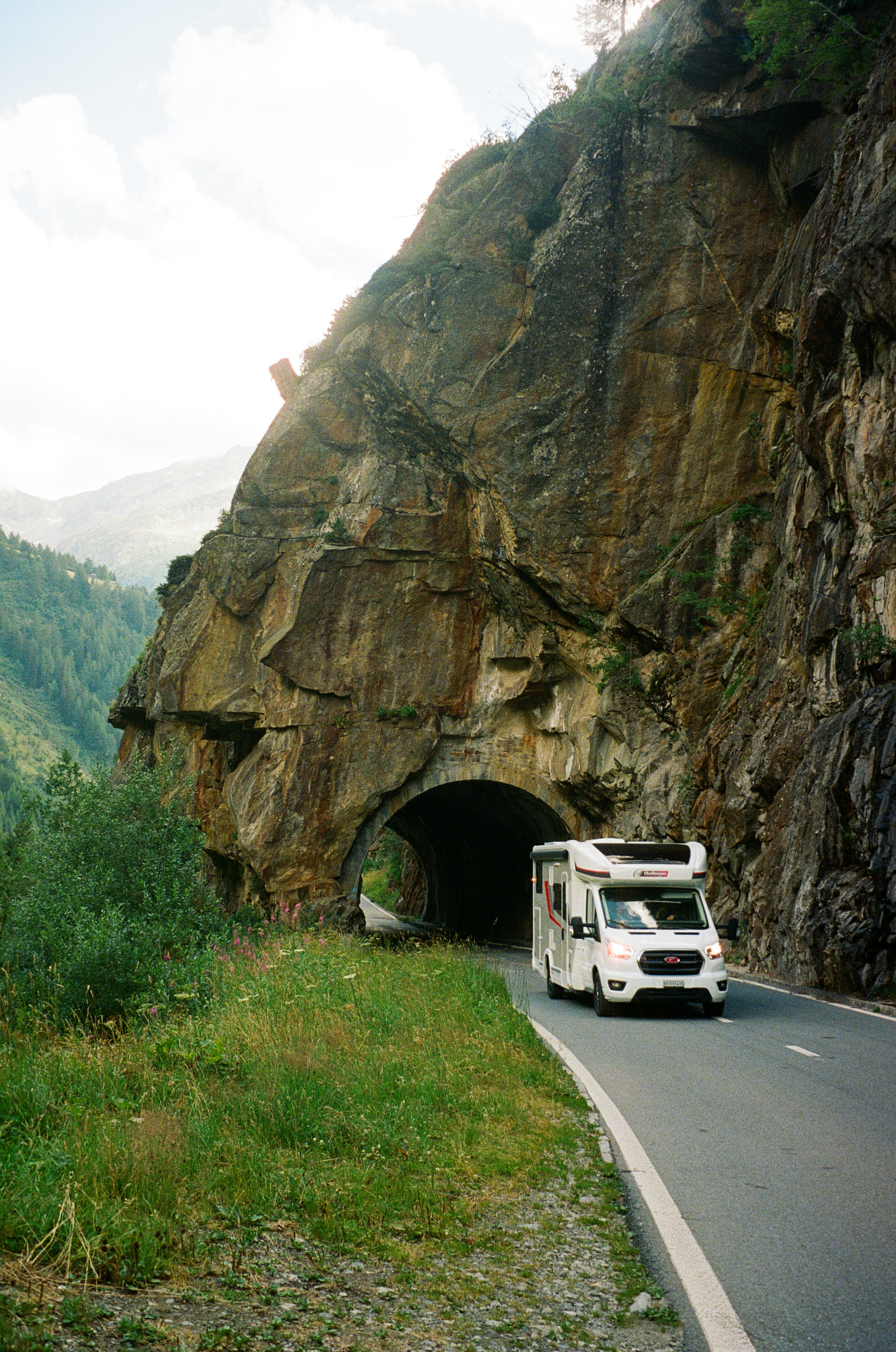 A white motorhome travels along a winding mountain road toward a dark tunnel carved into a granite cliff. Lush greenery lines the roadside as the scene captures travel through rugged terrain.