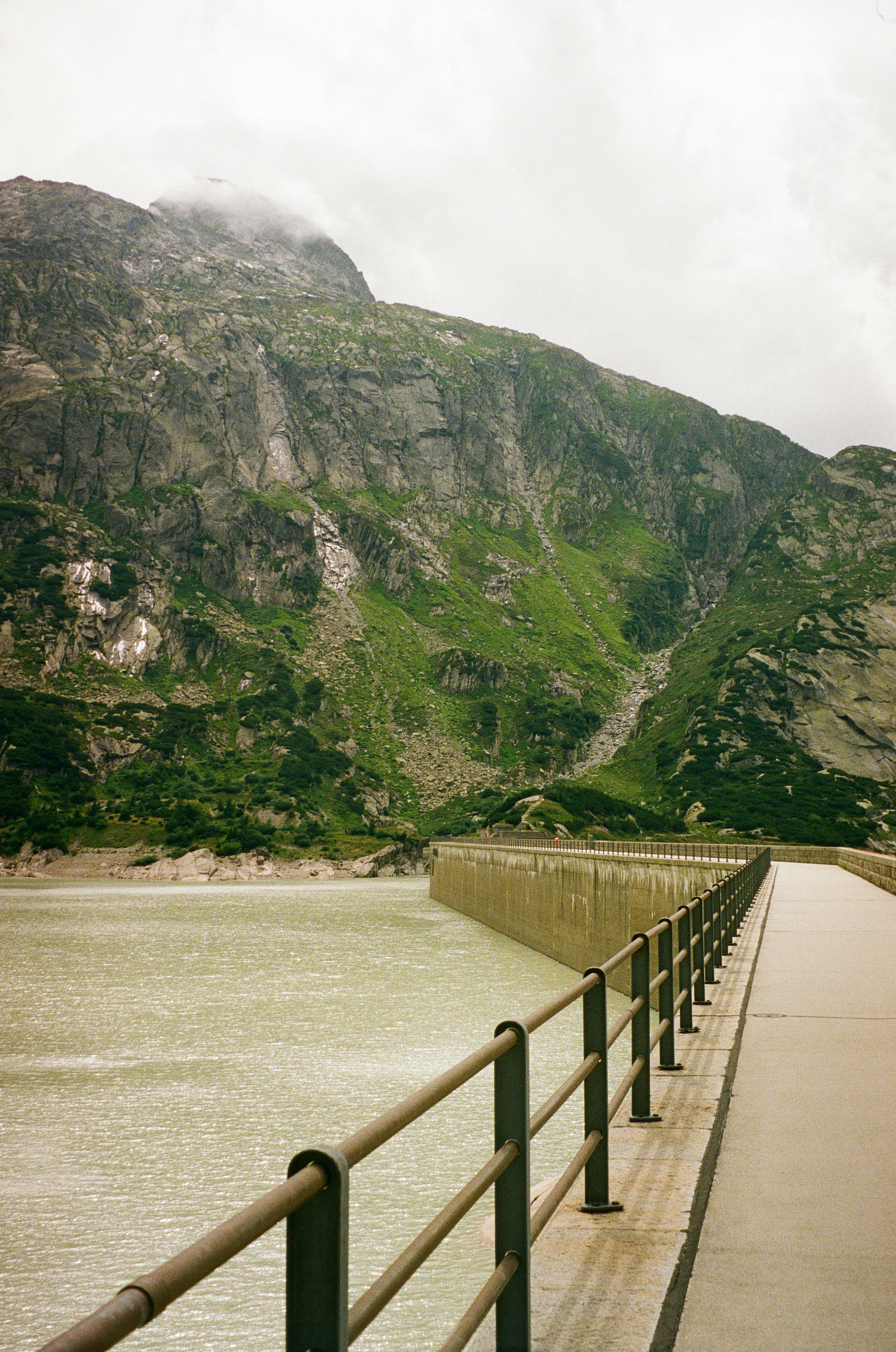 Photograph of a concrete pier extending toward emerald cliffs under an overcast sky. The railing guides the eye toward distant green mountains.