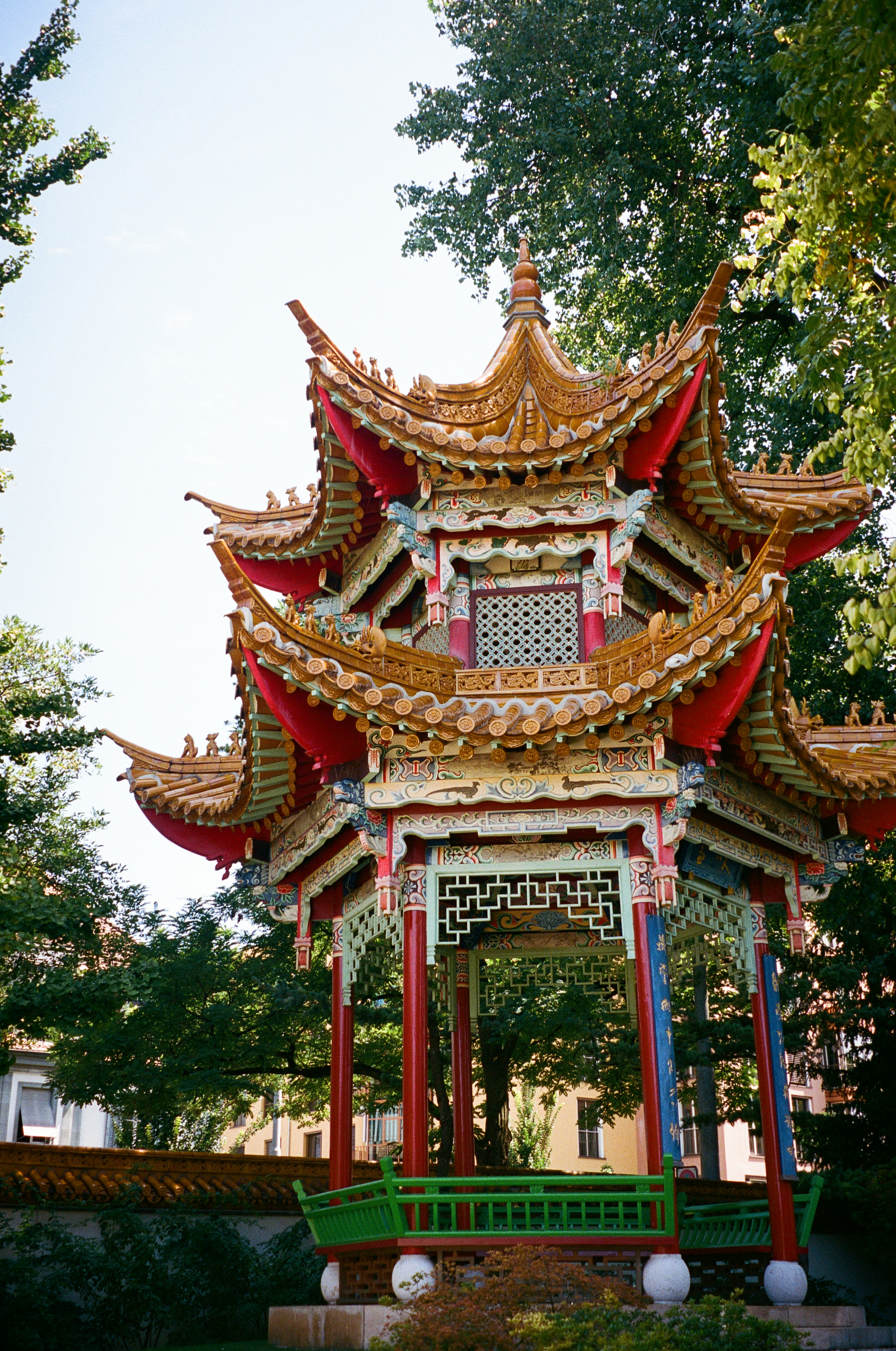 Colorful, multi-tier Chinese pavilion framed by lush trees, with intricate eaves and vibrant red, blue, and gold accents.