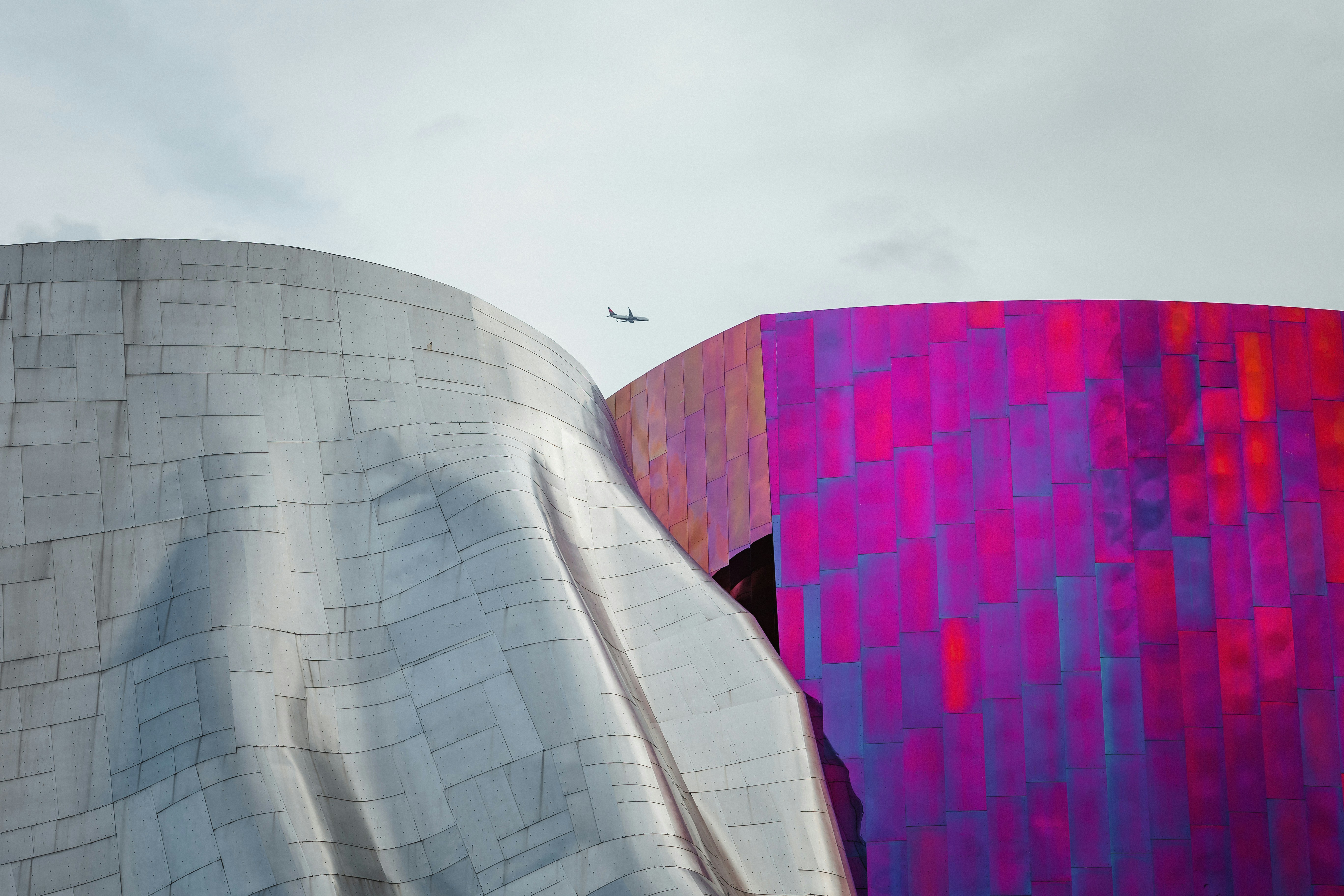 Plane glides over the seam where sculpted silver folds meet a magenta-tiled wall