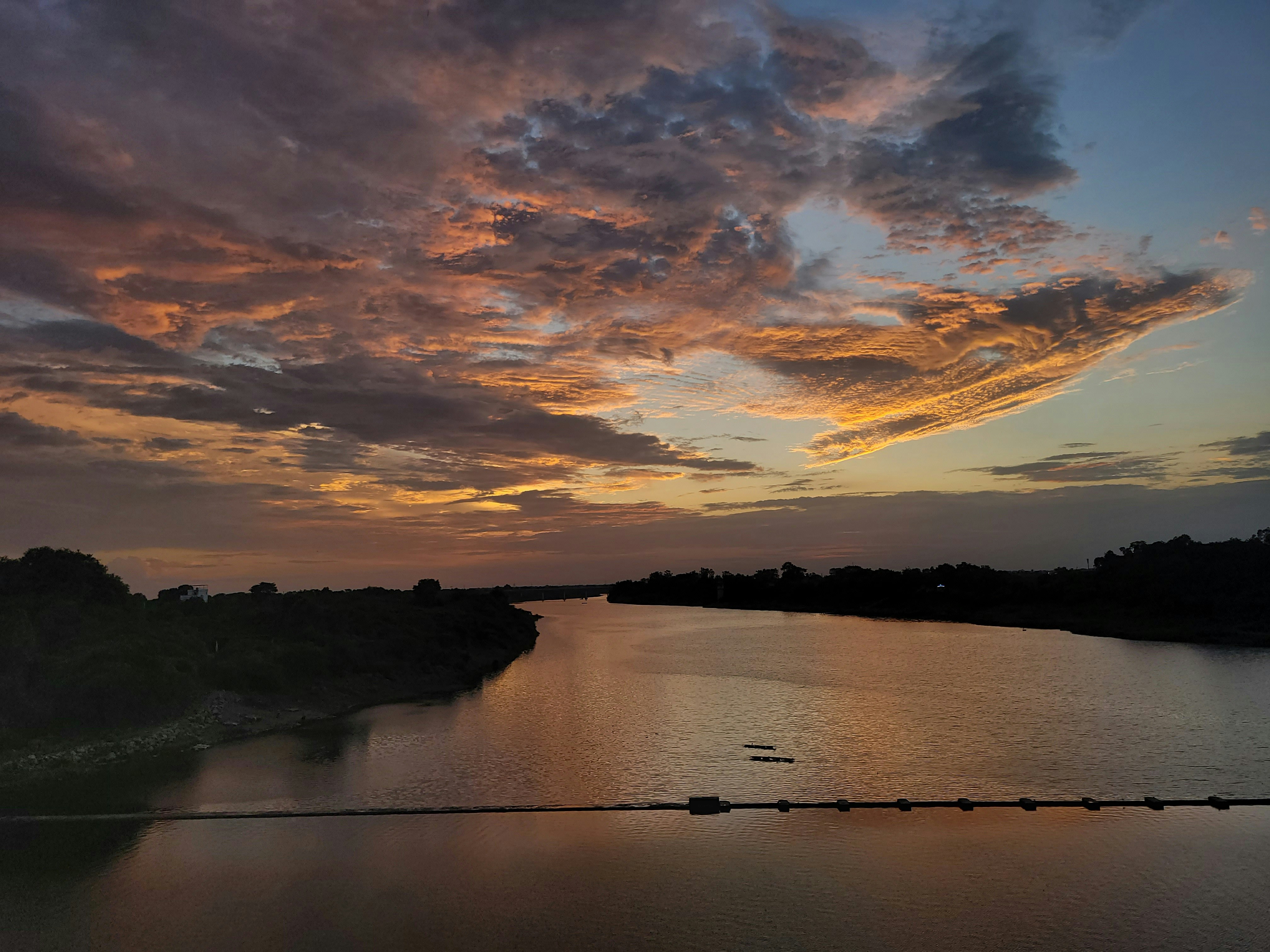 River beneath a dramatic sky at sunset with vibrant clouds and silhouettes of distant trees.