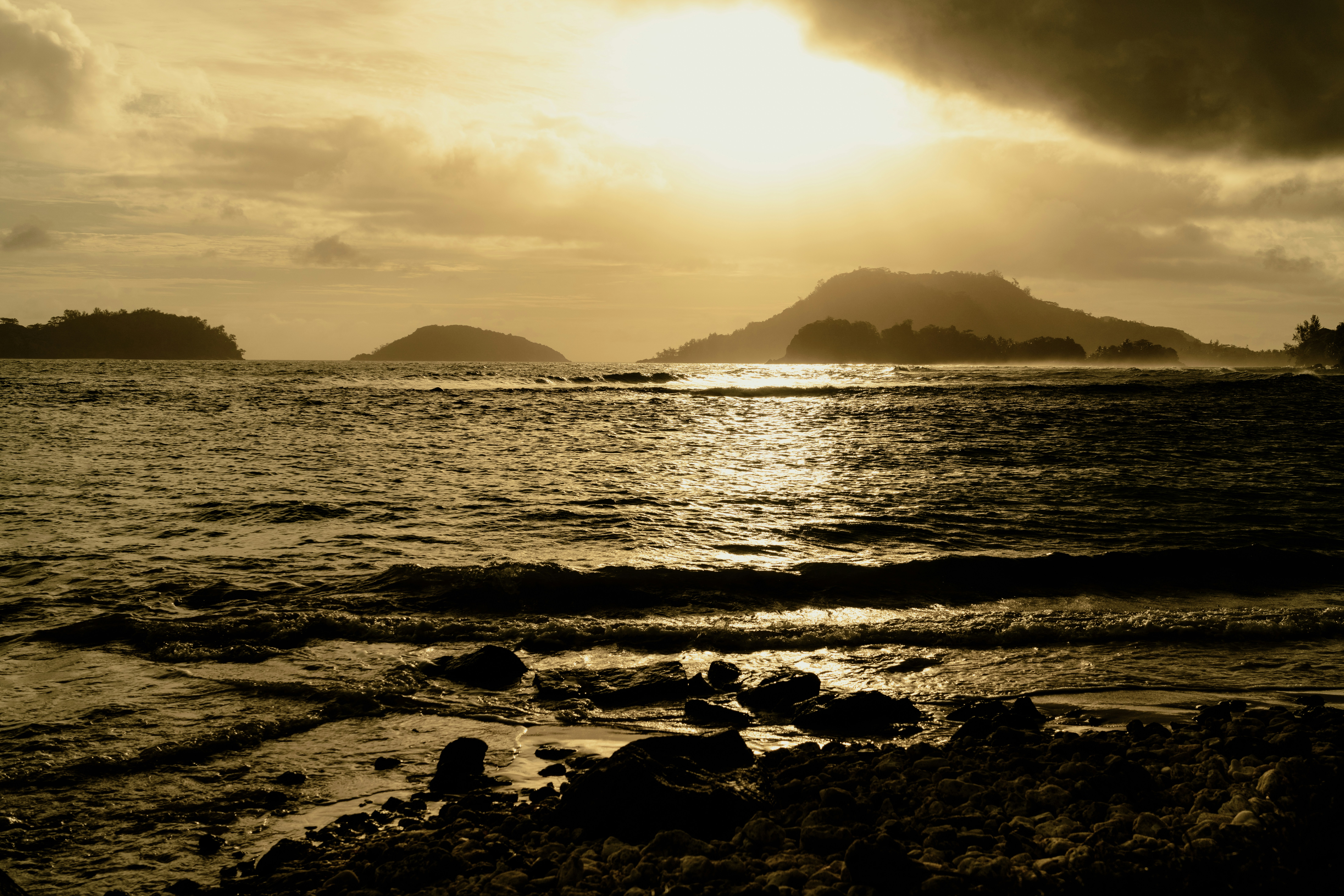 Sunset over a rocky shoreline with islands silhouetted in the distance and dramatic clouds overhead.