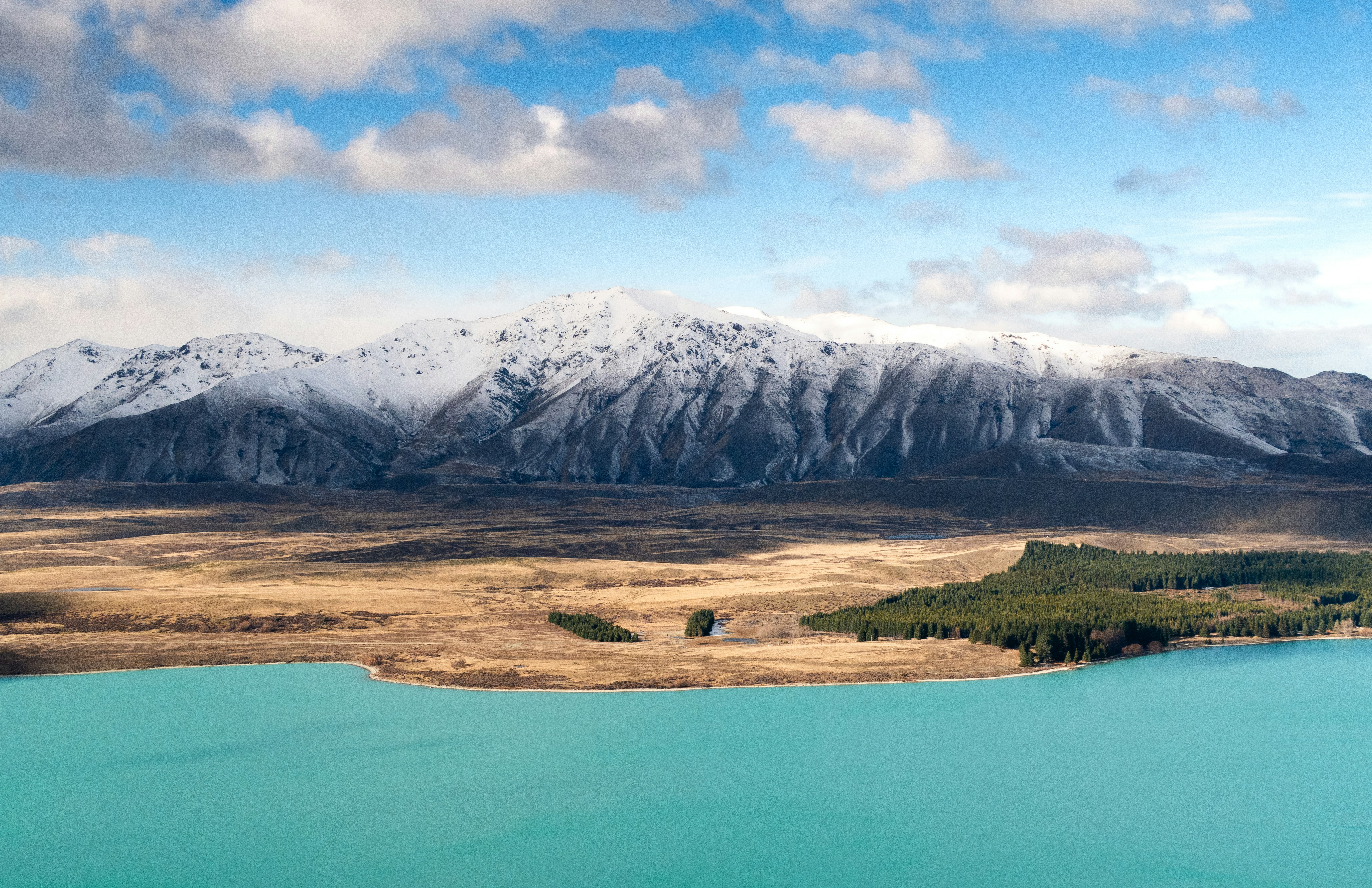 Turquoise lake laps a sandy shoreline, with a pine peninsula extending toward a snow-dusted mountain range. This landscape photograph emphasizes color contrast and expansive depth.