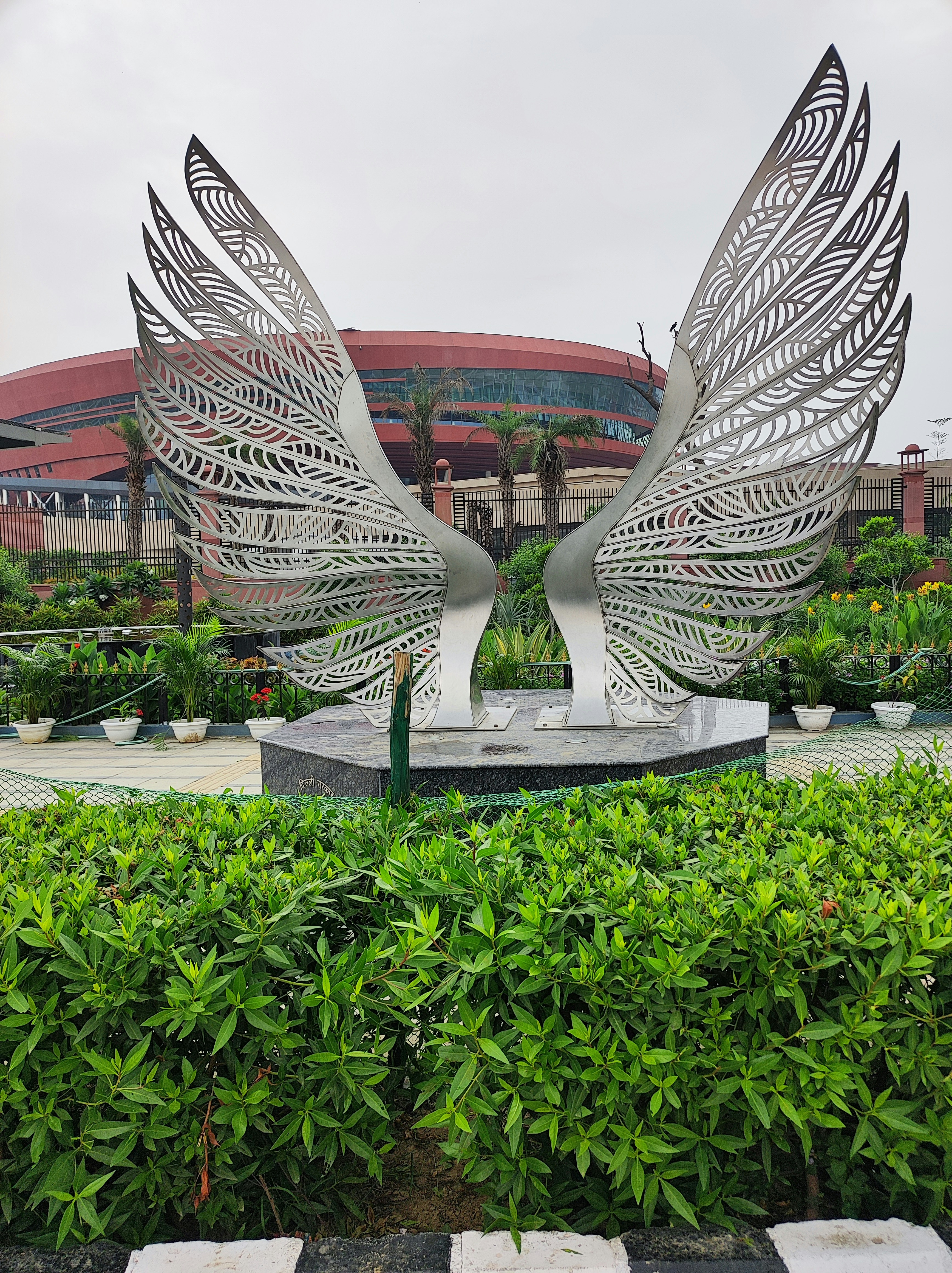 Winged stainless sculpture: mirrored arcs rising over manicured hedges