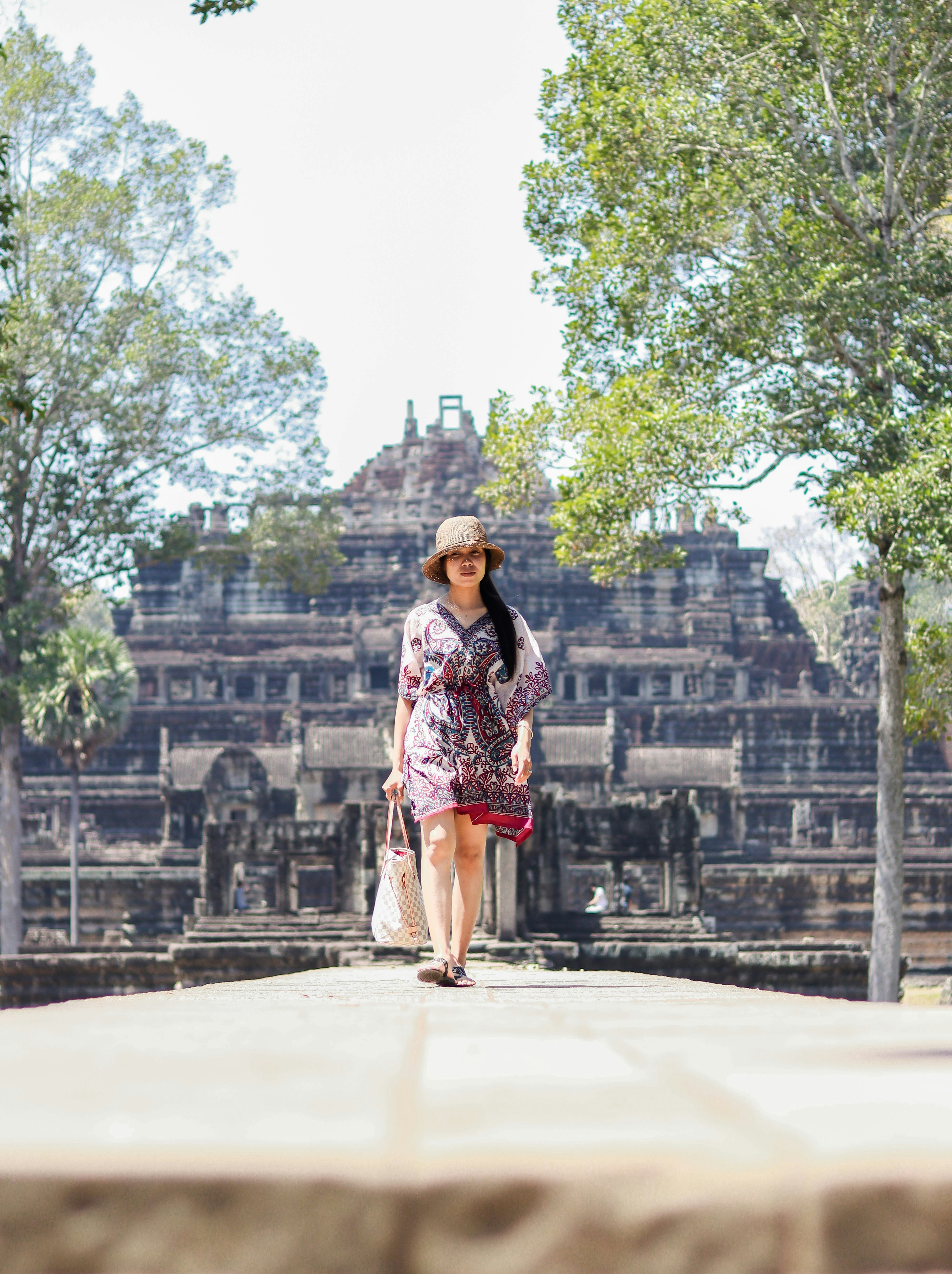 A woman walks confidently along a stone pathway leading to a historic temple, framed by lush greenery and ancient architecture.