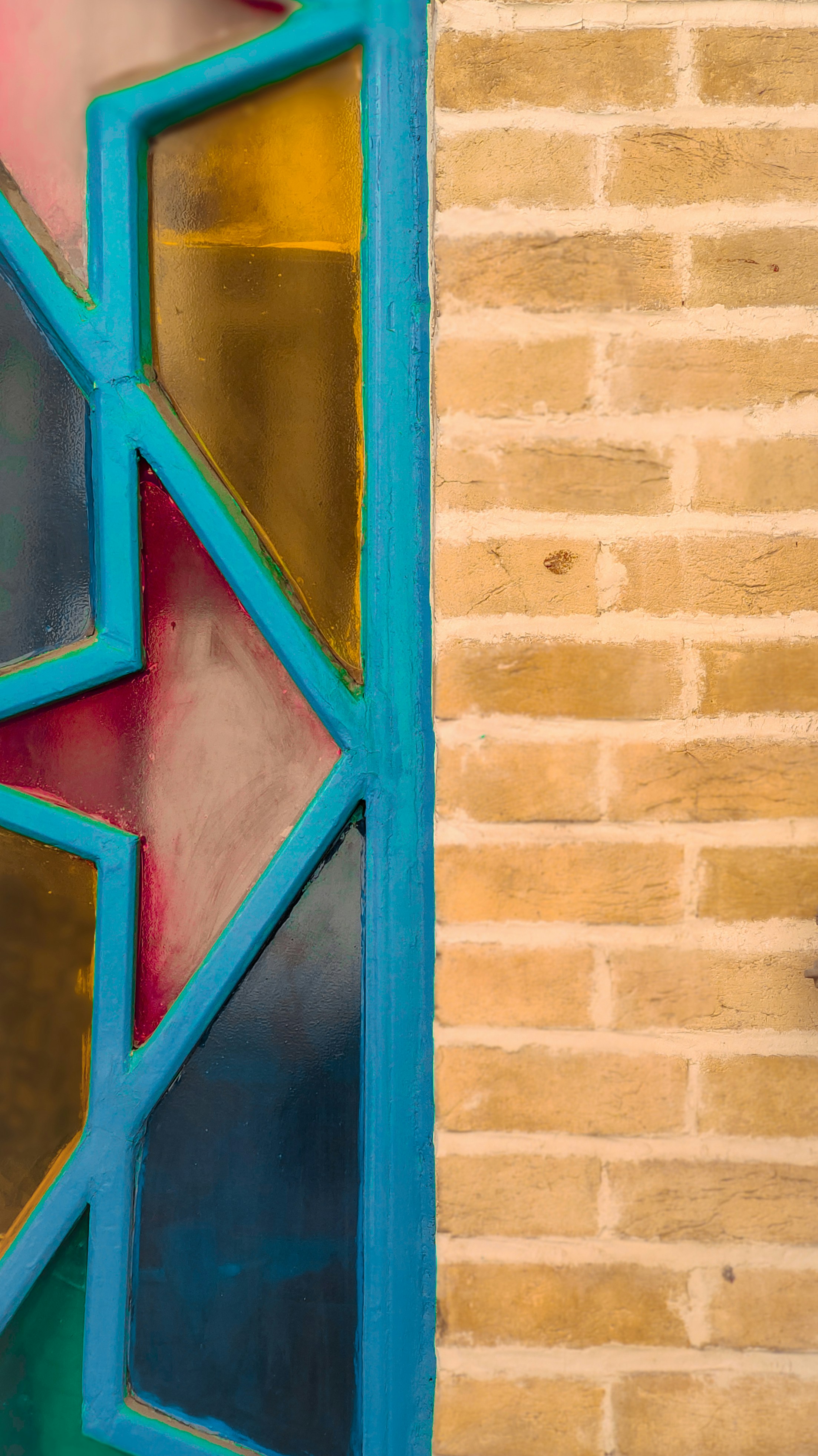 Geometric stained-glass panel with a vivid turquoise frame sits beside a warm brick wall. The colorful panes create a bold contrast with the brickwork.