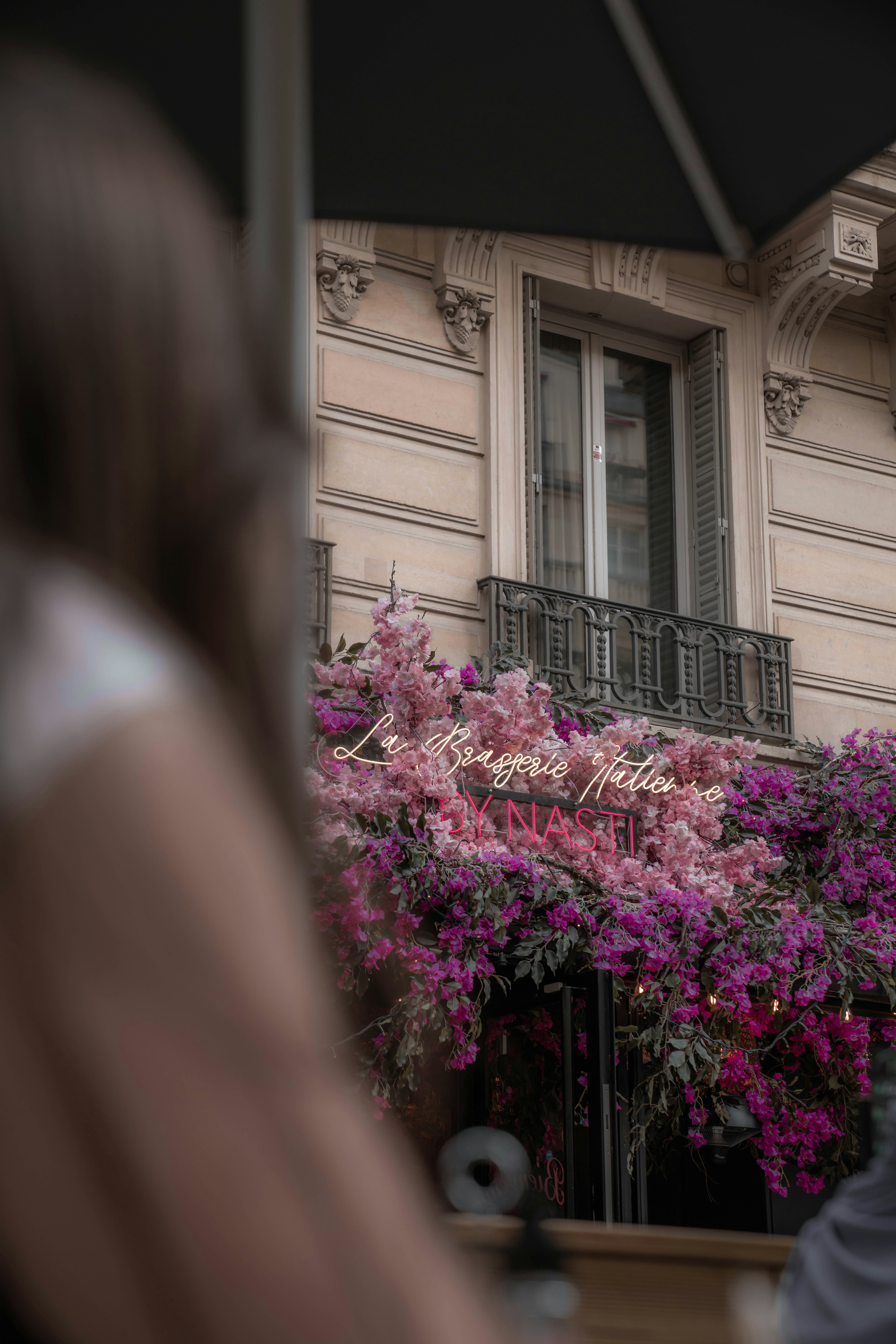 Parisian café storefront framed by pink bougainvillea around a wrought-iron balcony and a cursive neon sign.
