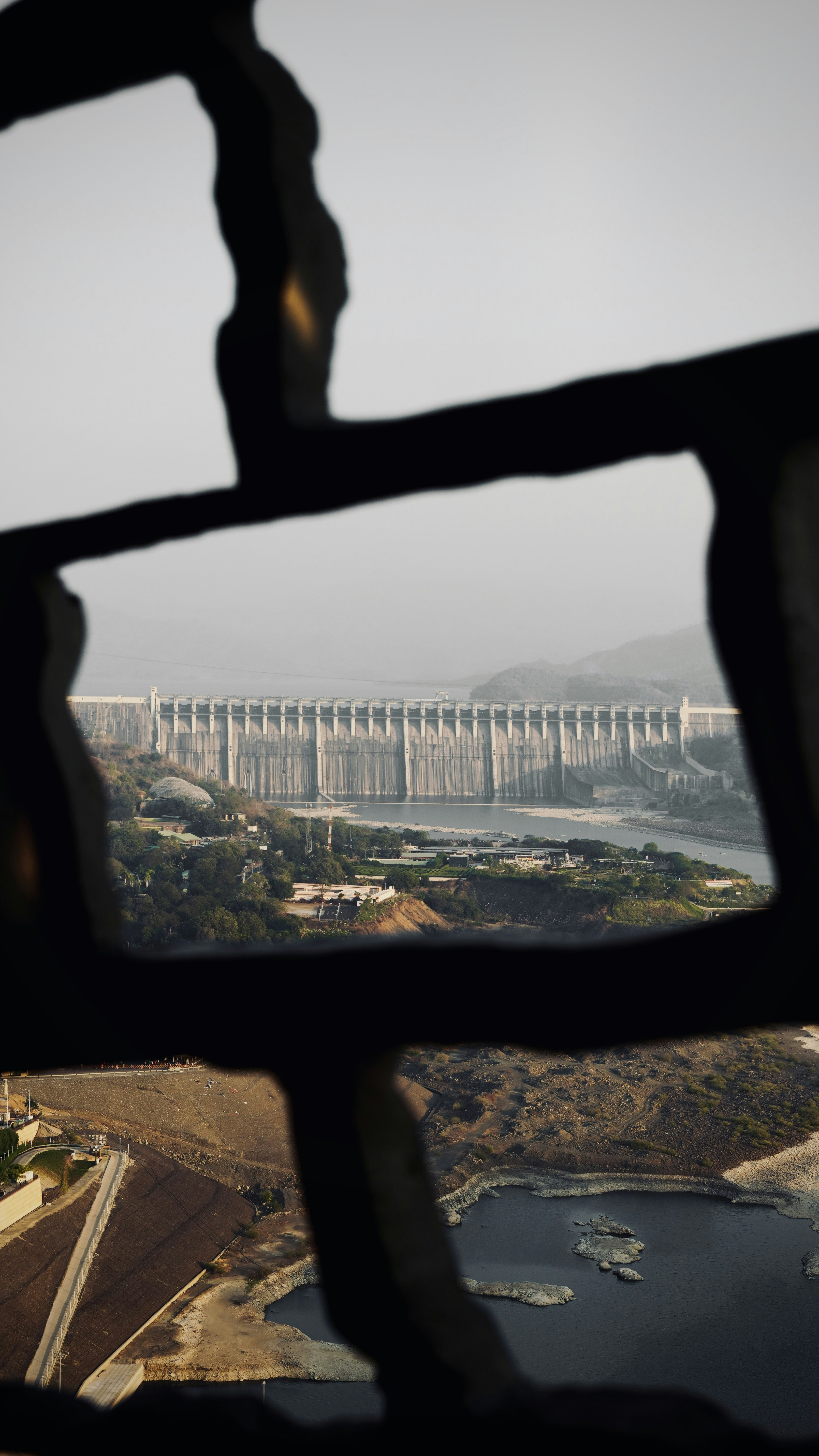 A dam viewed through a textured frame, showcasing its structure against a hazy landscape. The scene emphasizes the interplay between nature and human engineering.