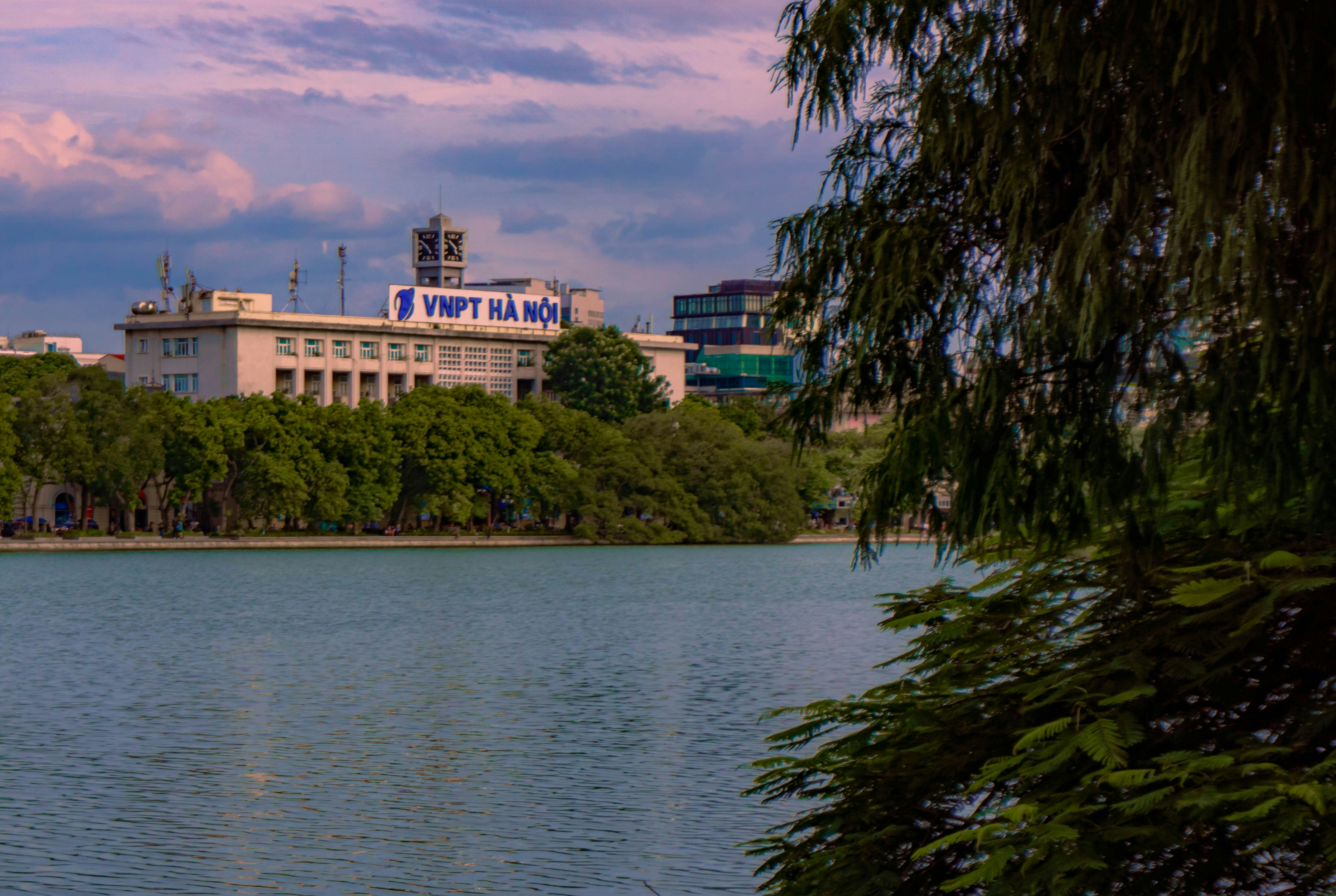 Calm water reflecting a skyline of buildings under a vibrant sky, framed by lush foliage.