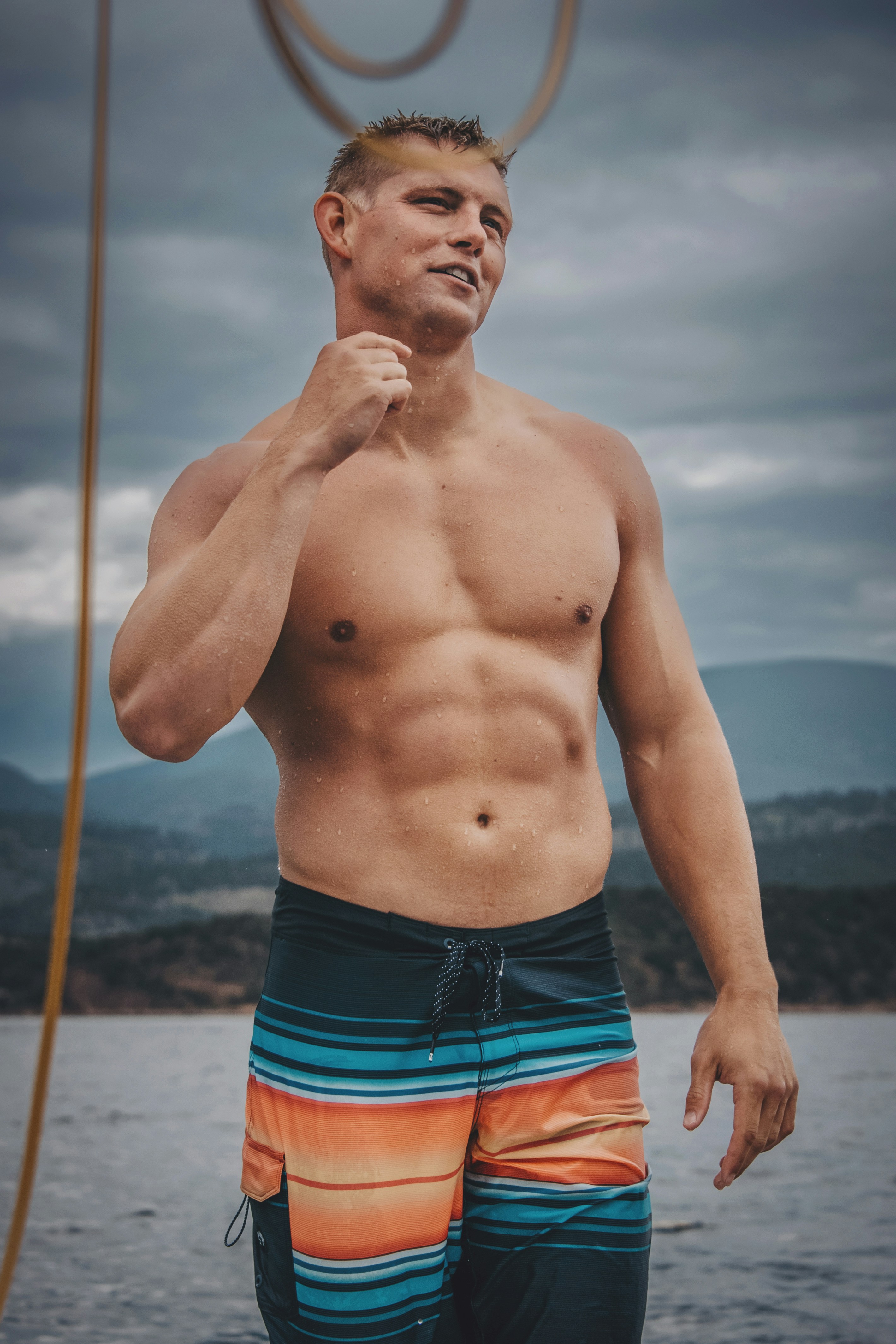 A muscular man poses confidently by the water, showcasing vibrant swim trunks against a backdrop of moody skies and distant hills.