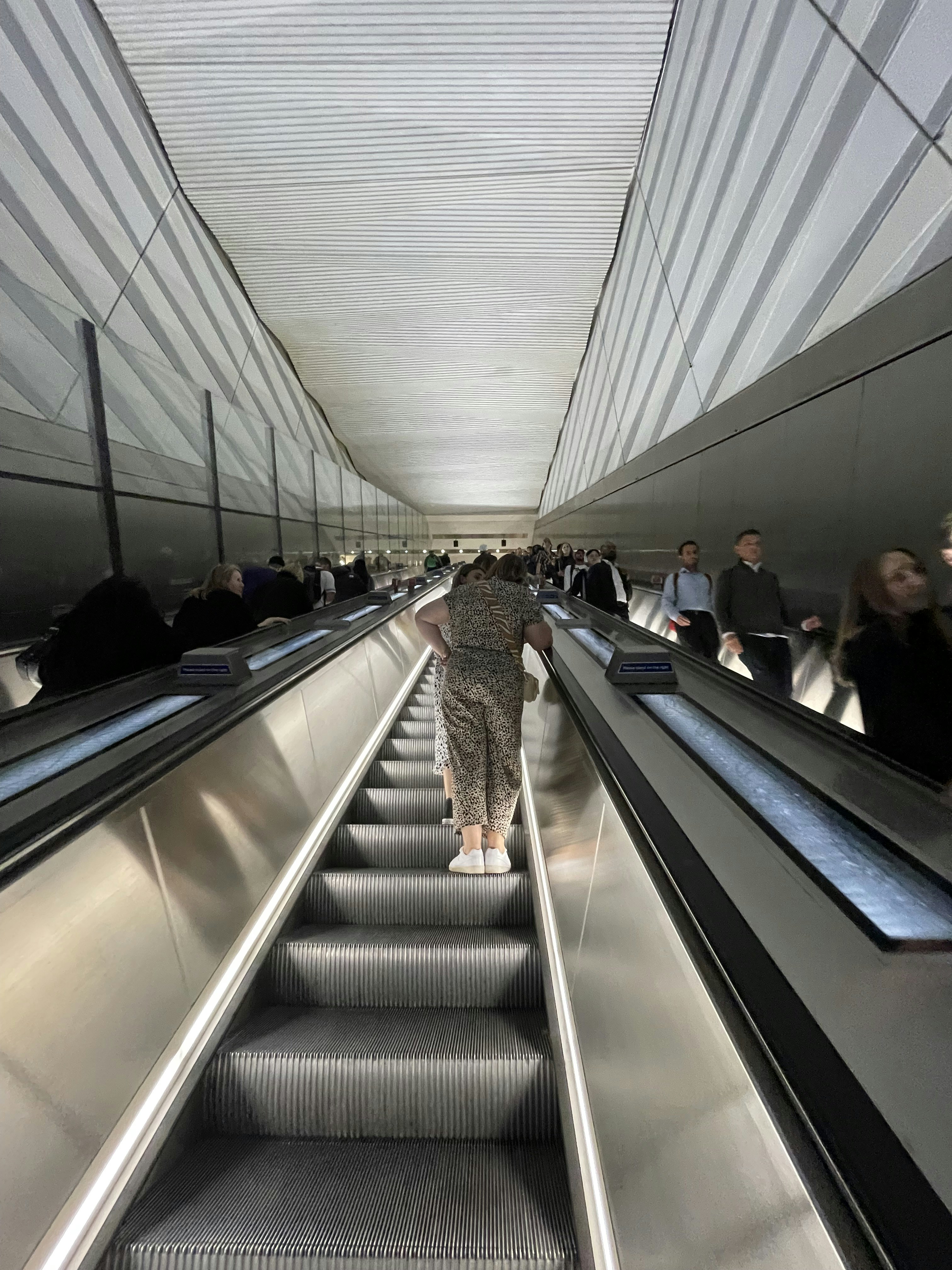 Escalator ascending in a modern architectural space, surrounded by a flow of commuters. The sleek lines and reflective surfaces create a dynamic visual experience.