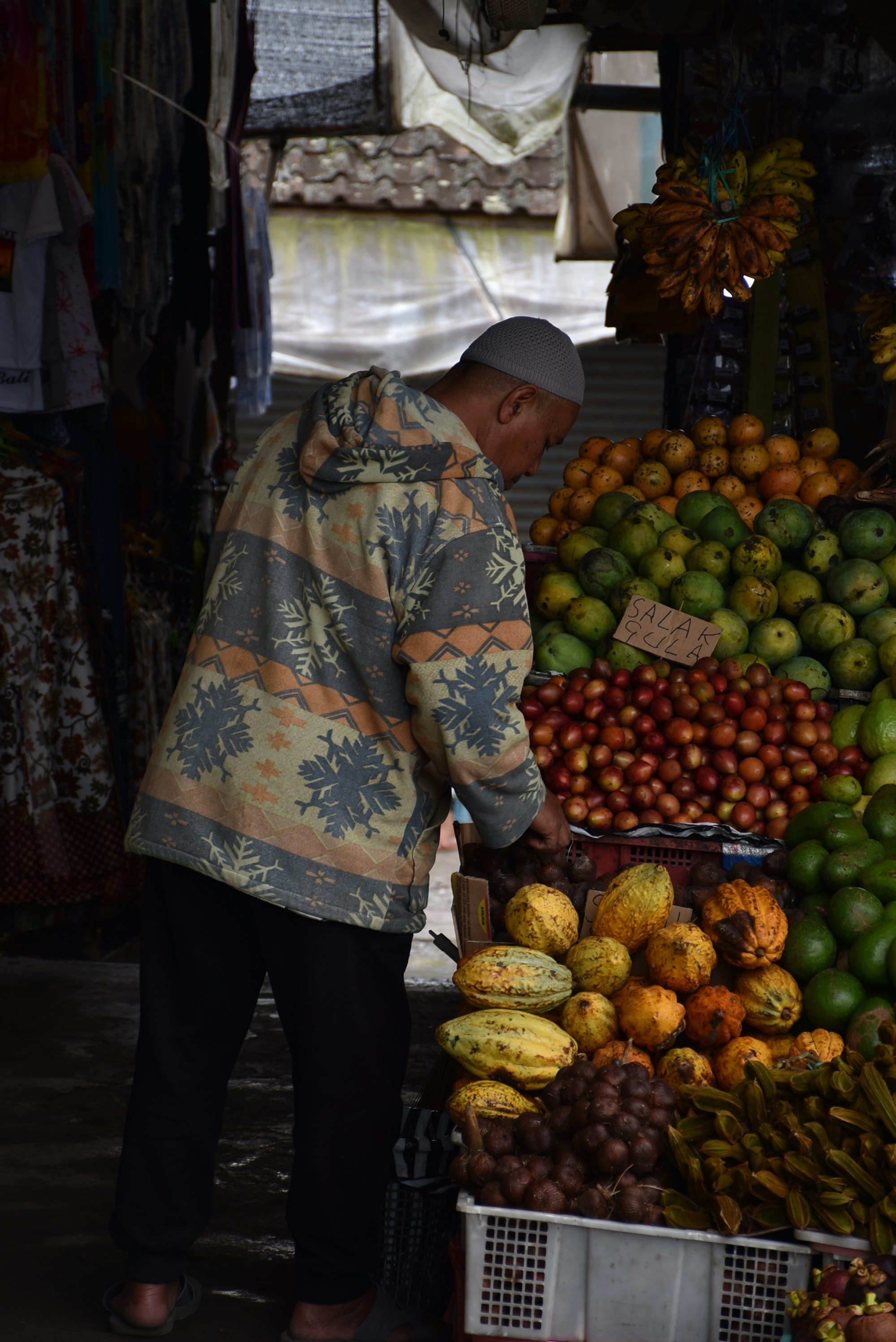 Shadowed Stall, Colorful Harvest: A shopper weighs fruit beneath a dim market canopy