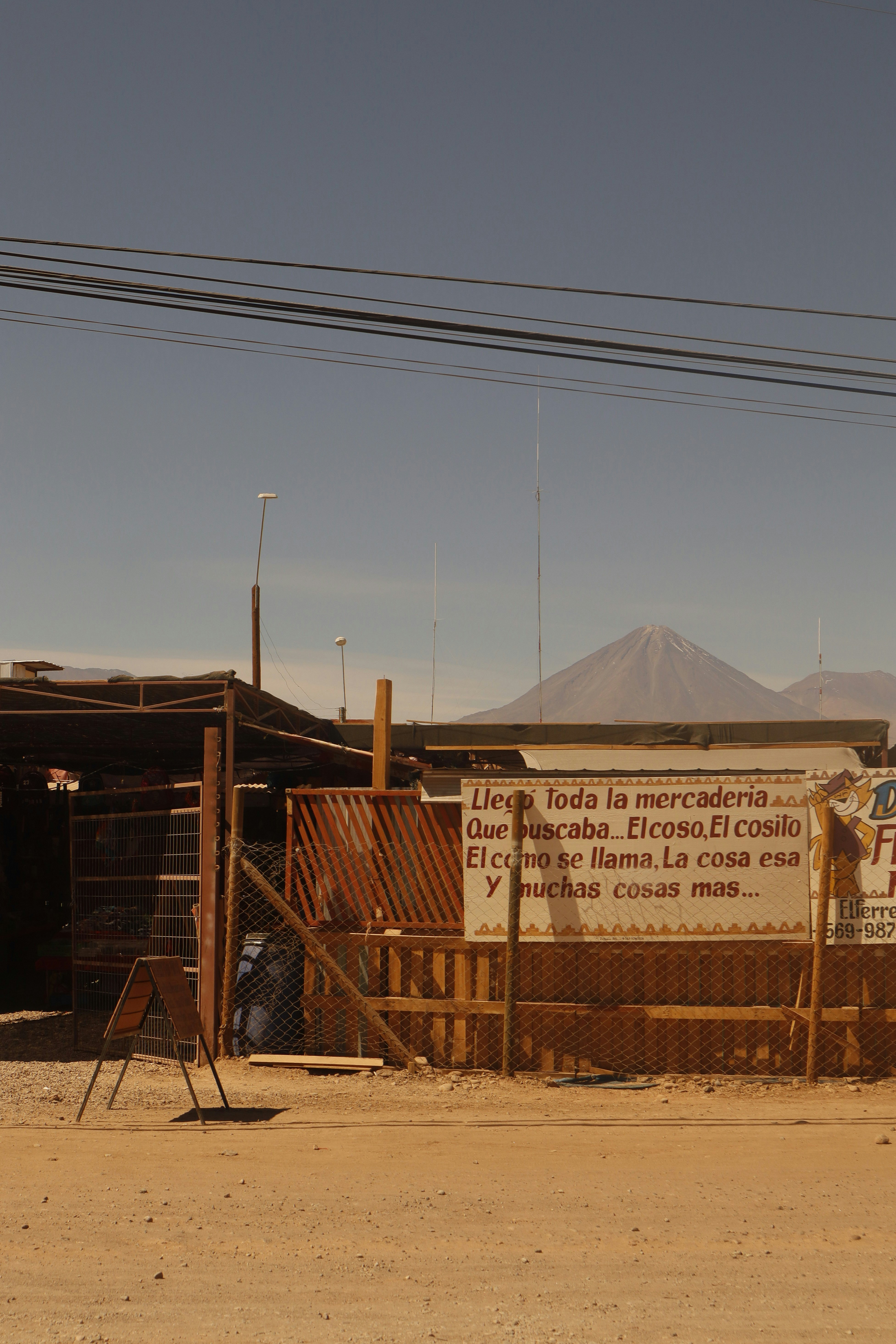 Dusty Roadside Market Framed by Fence and a Distant Volcano Horizon