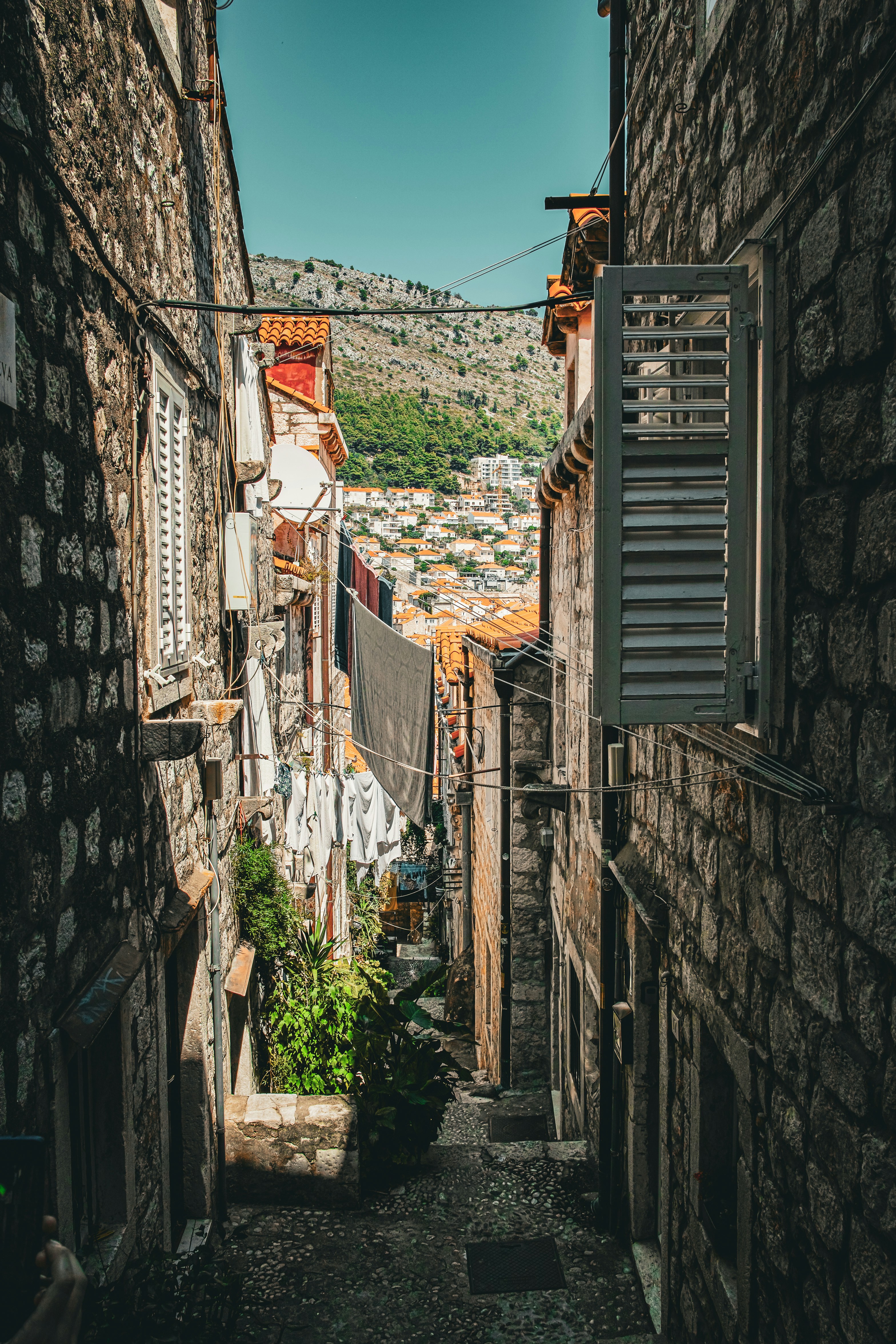 Narrow sunlit alley framed by ancient stone walls, with laundry lines and greenery, leads toward a sunlit hillside town in the distance.