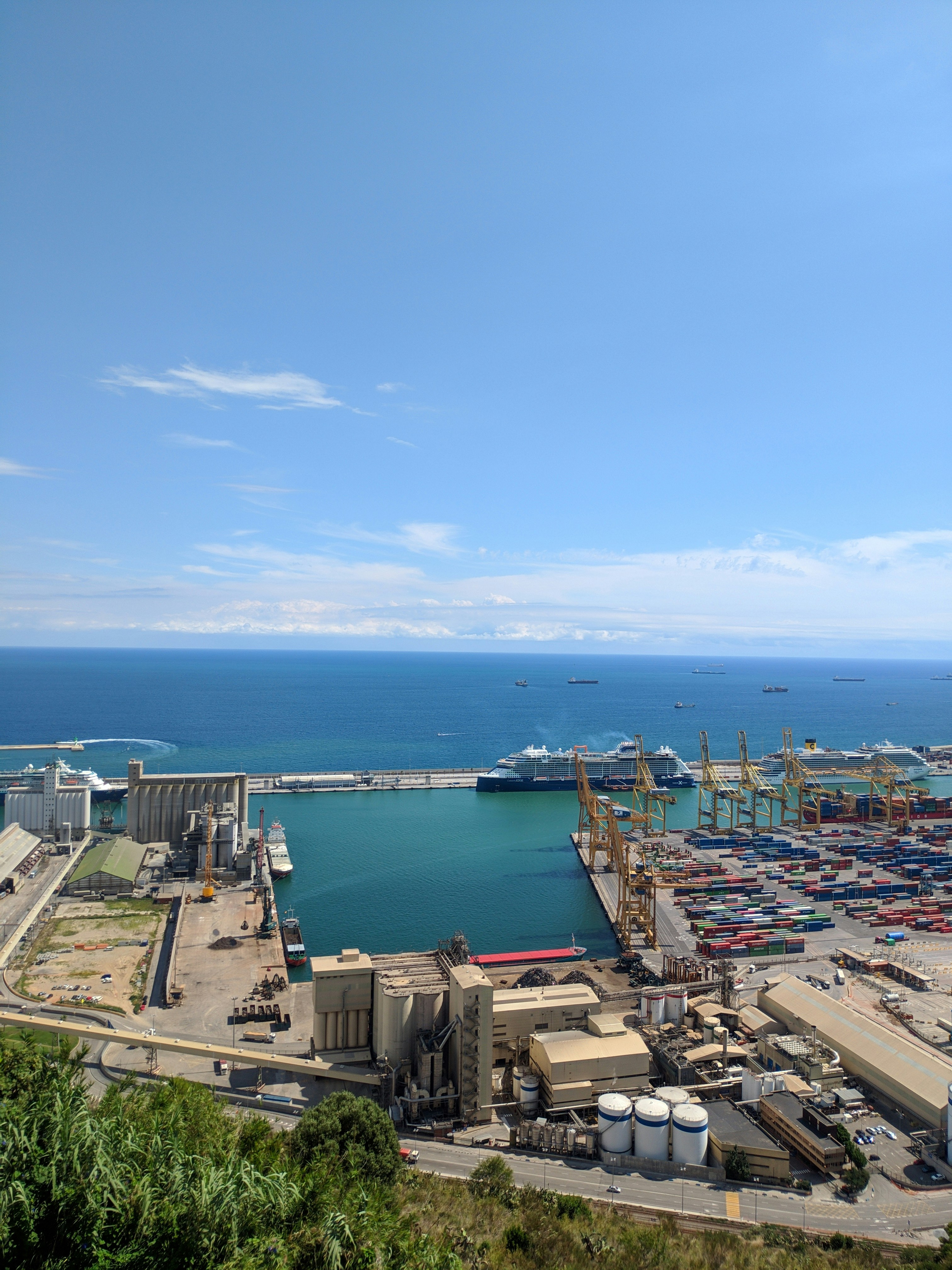 A port scene with rows of colorful containers, towering cranes, and a calm blue sea beneath a clear sky.