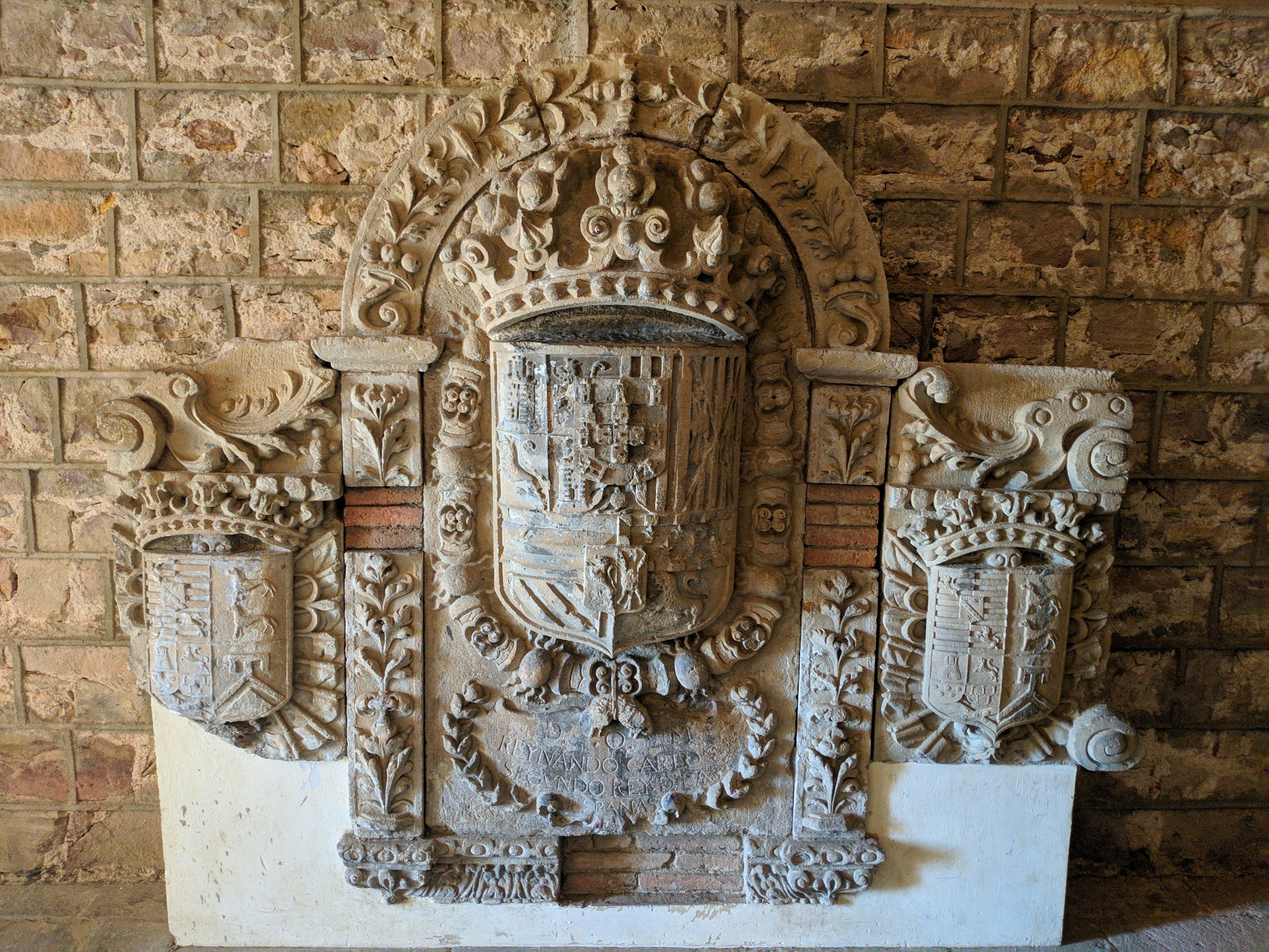 Intricately carved heraldic crest featuring a central crowned shield flanked by two smaller shields, set into a weathered brick wall.