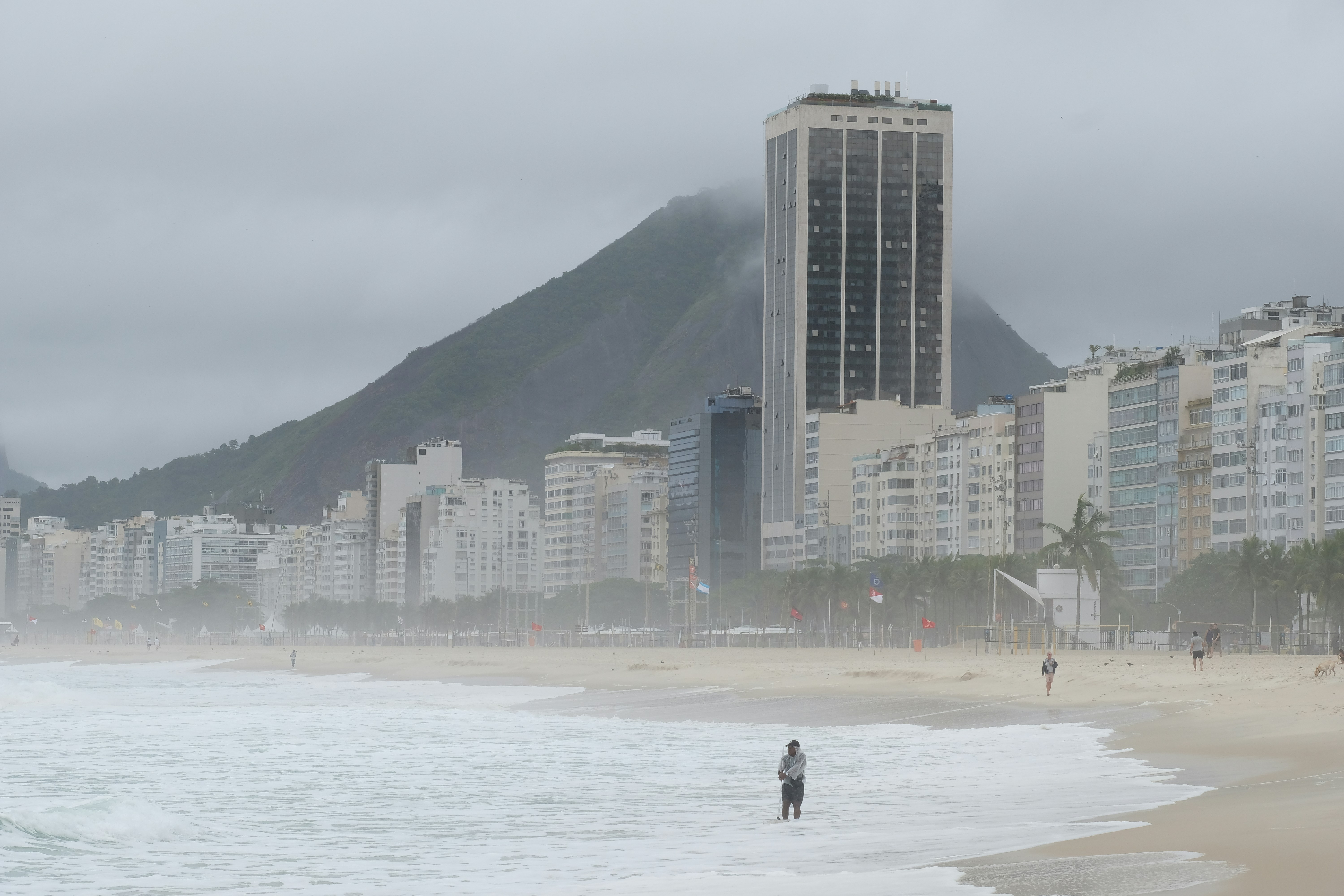 Person walking along a cloudy beach with a city skyline and mountain backdrop.