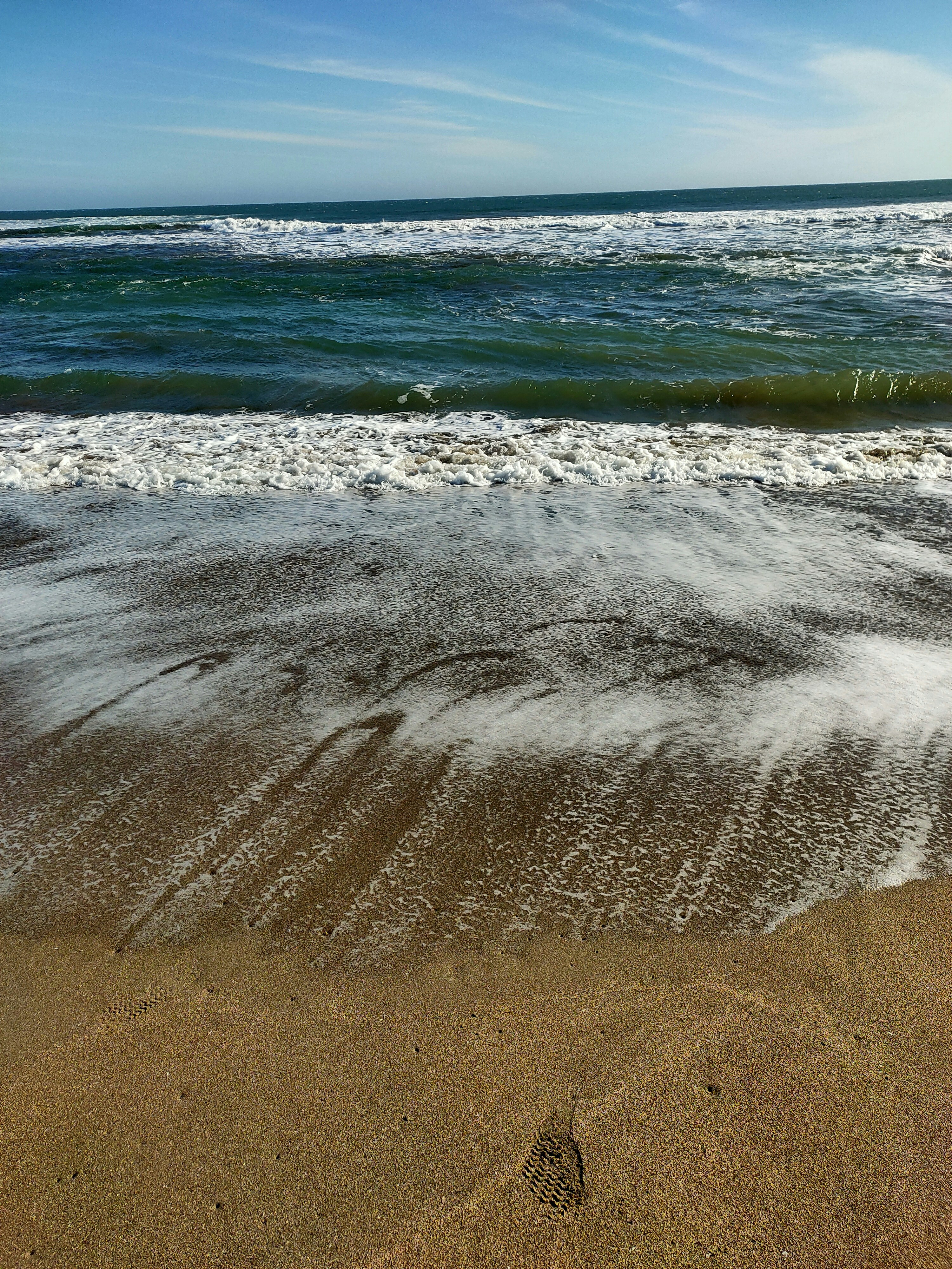 Foamy shoreline washes over smooth sand as green-blue waves roll in under a clear sky.