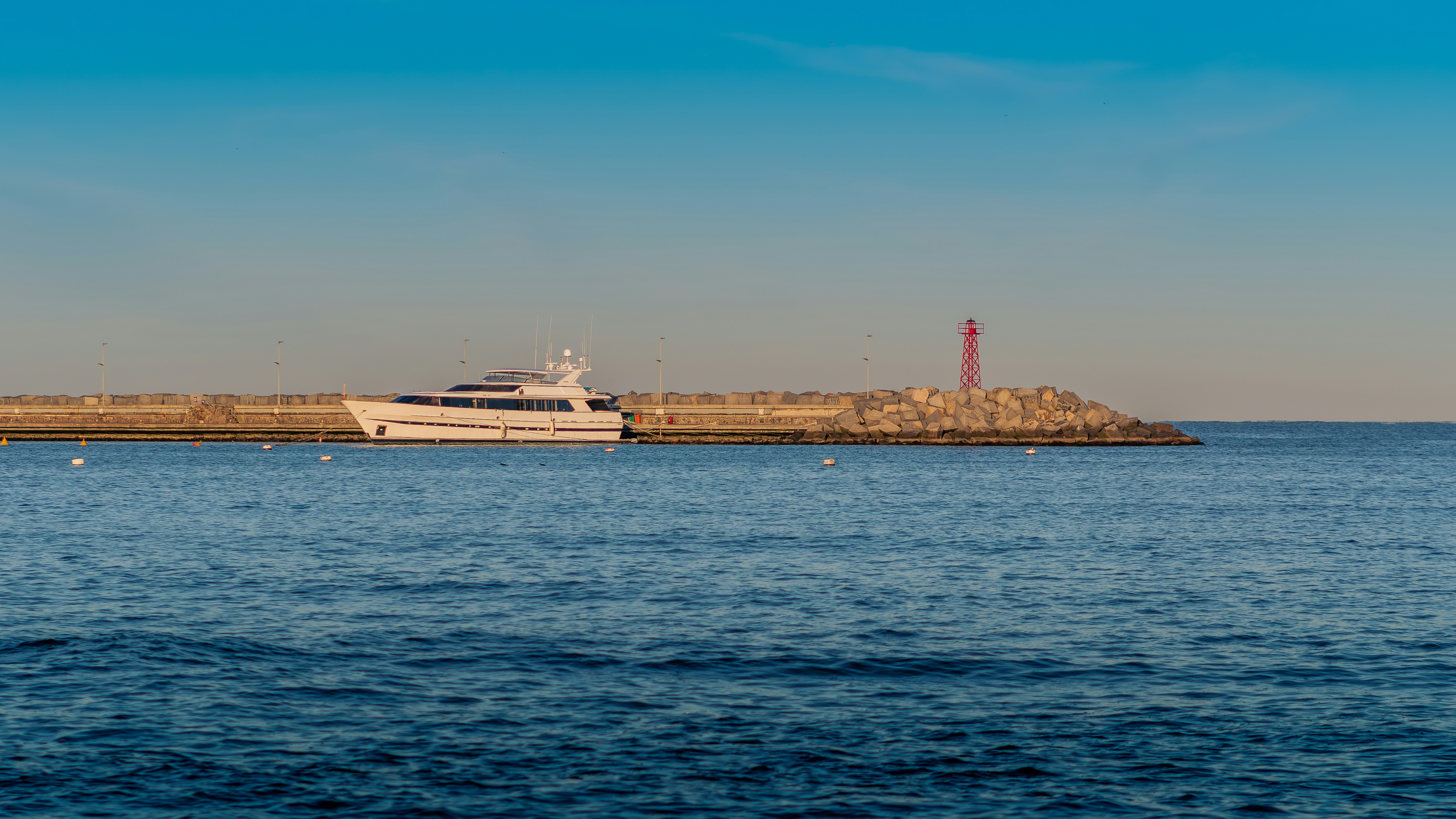 Large yacht docked near a rocky breakwater with a red beacon under a clear blue sky.