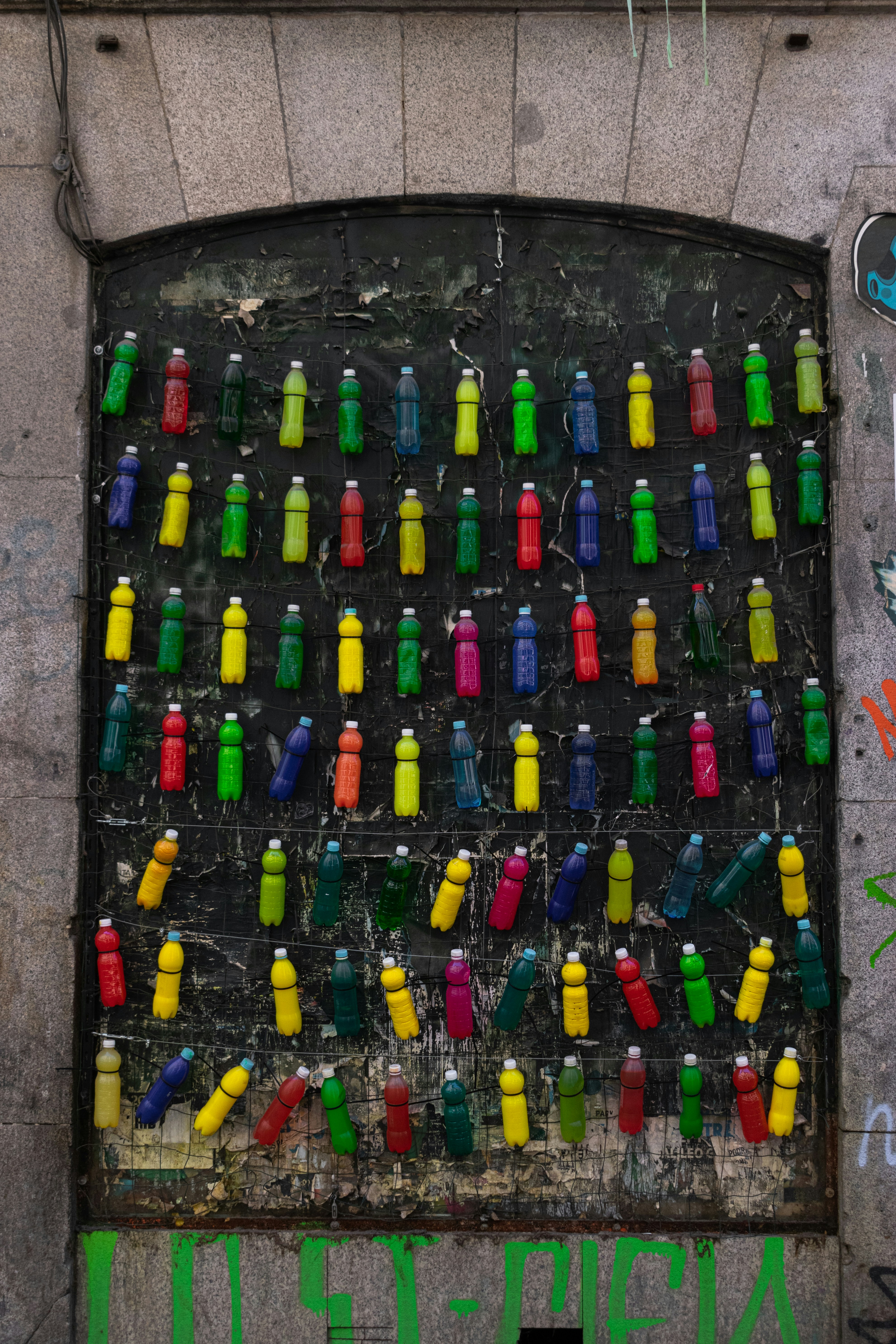 Rainbow Grid of Bottles on a Weathered Urban Gate