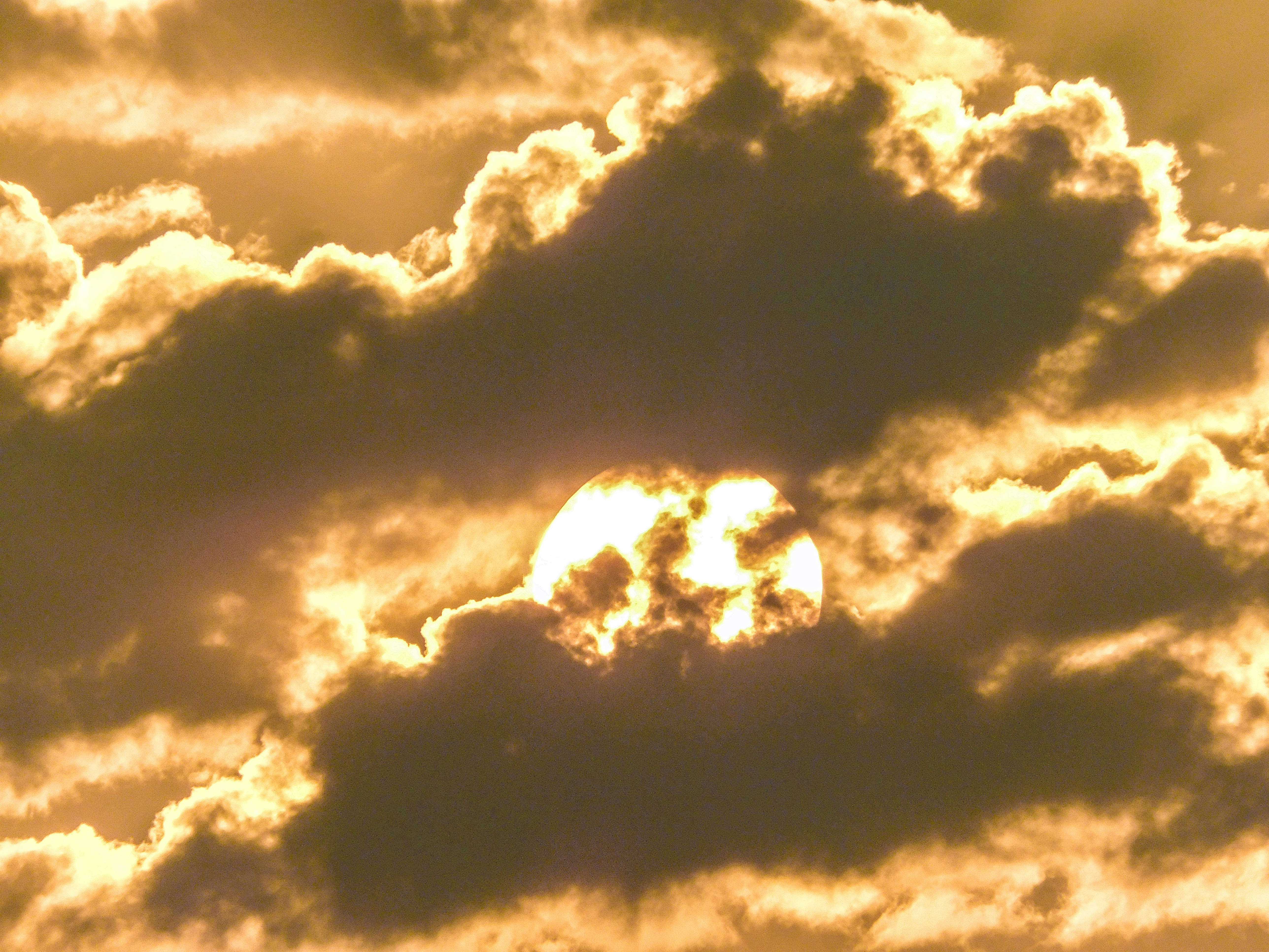 Golden-hour photograph of the sun peeking through ember-colored clouds, casting warm light across the sky. The scene emphasizes dramatic cloud edges and a quiet horizon.