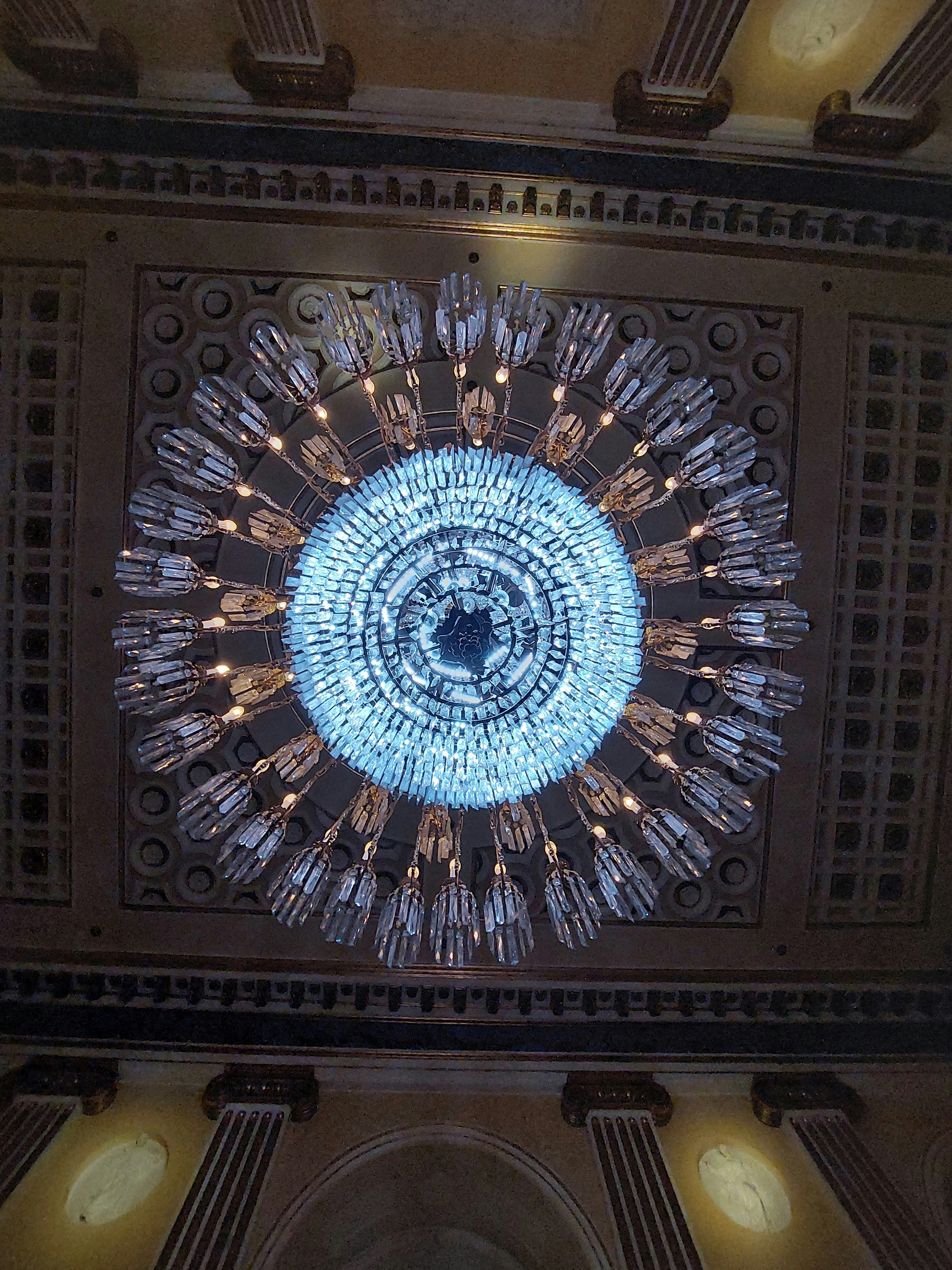 Circular crystal chandelier viewed from below, bathed in cool blue light against an ornate ceiling.