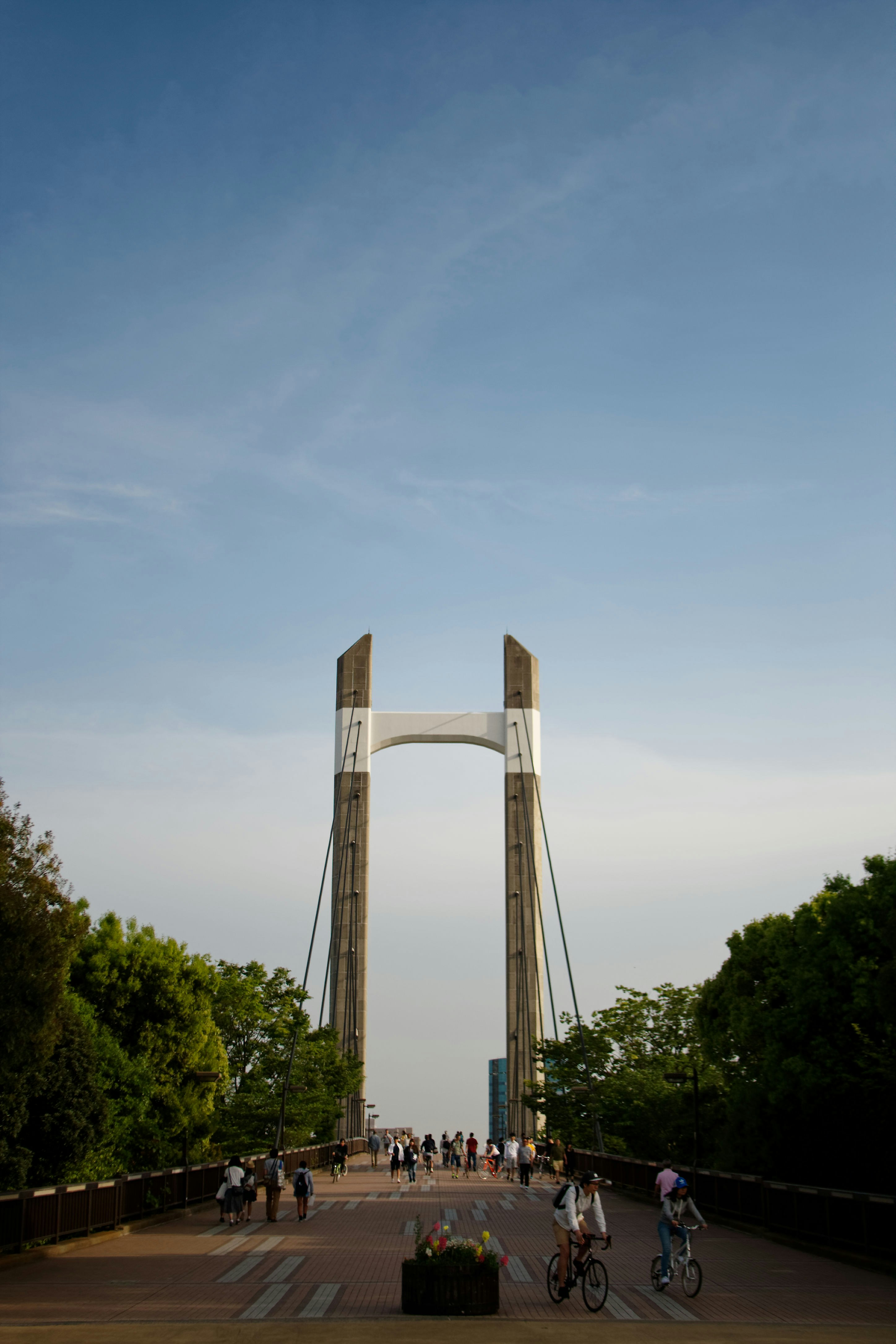 Photograph of pedestrians and cyclists on a walkway beneath a tall suspension arch, flanked by trees and a clear blue sky.