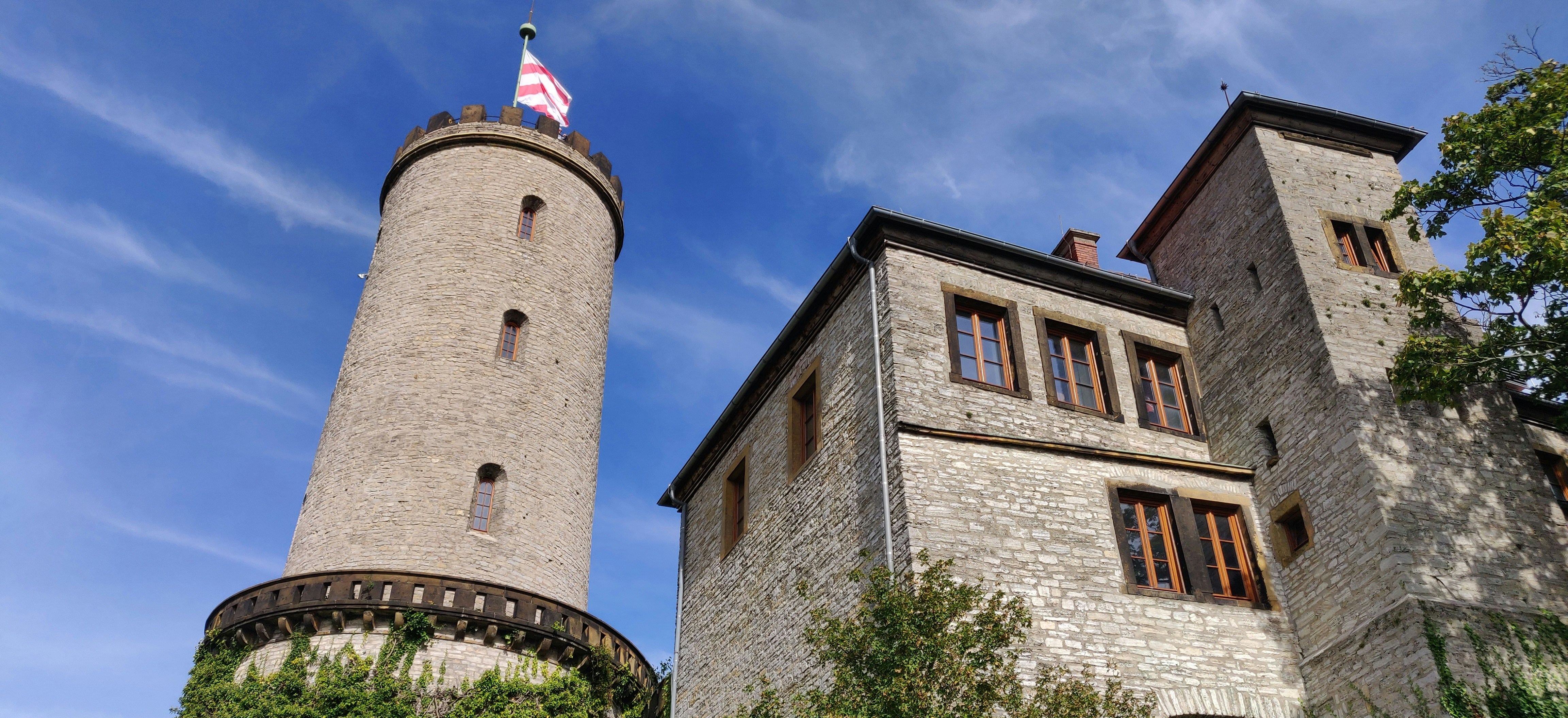 Stone tower with an American flag, adjacent to a historic building under a clear blue sky.