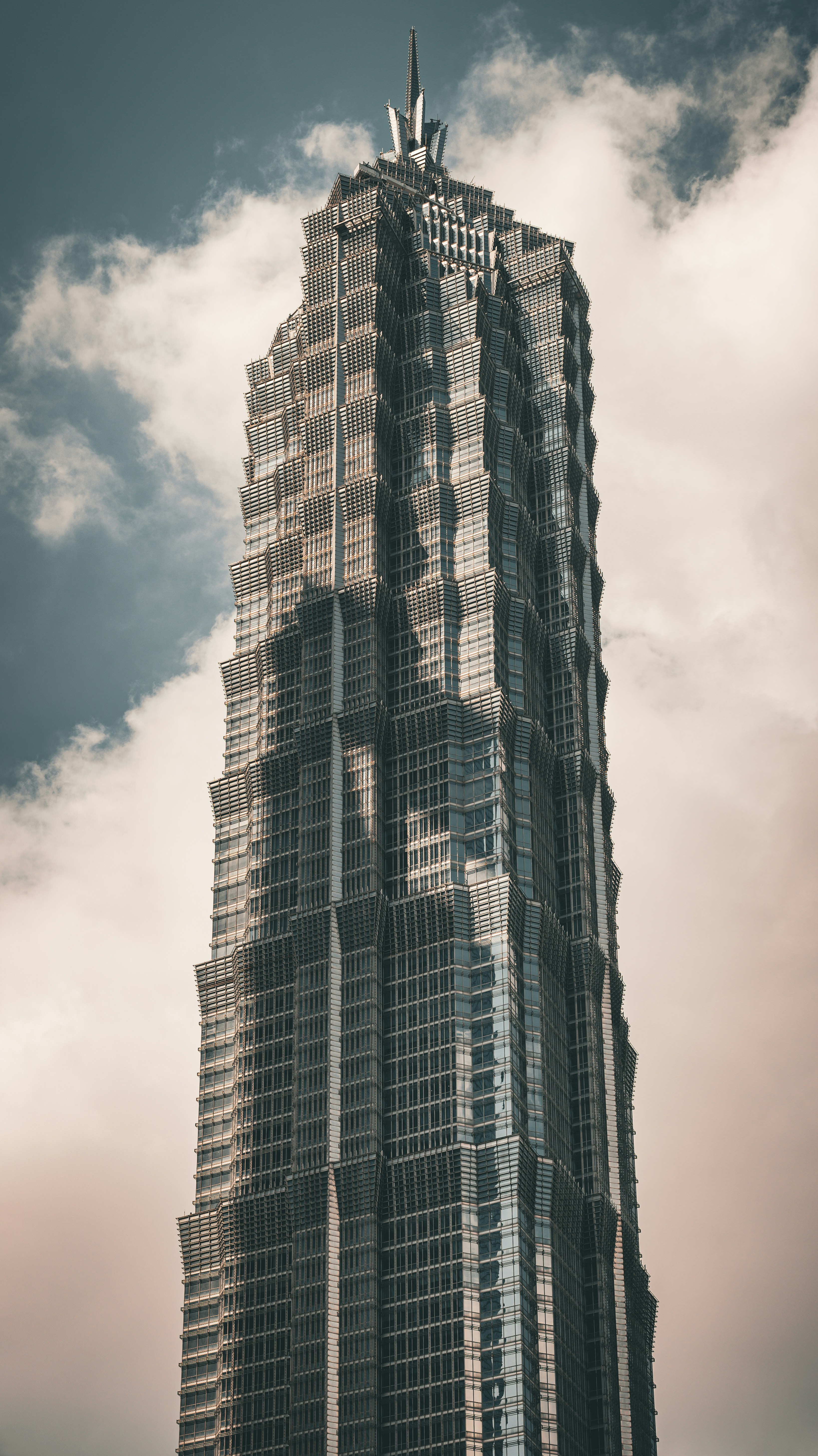 Photograph of a towering glass-and-metal skyscraper with a serrated, grid-like façade rising into a cloudy sky.