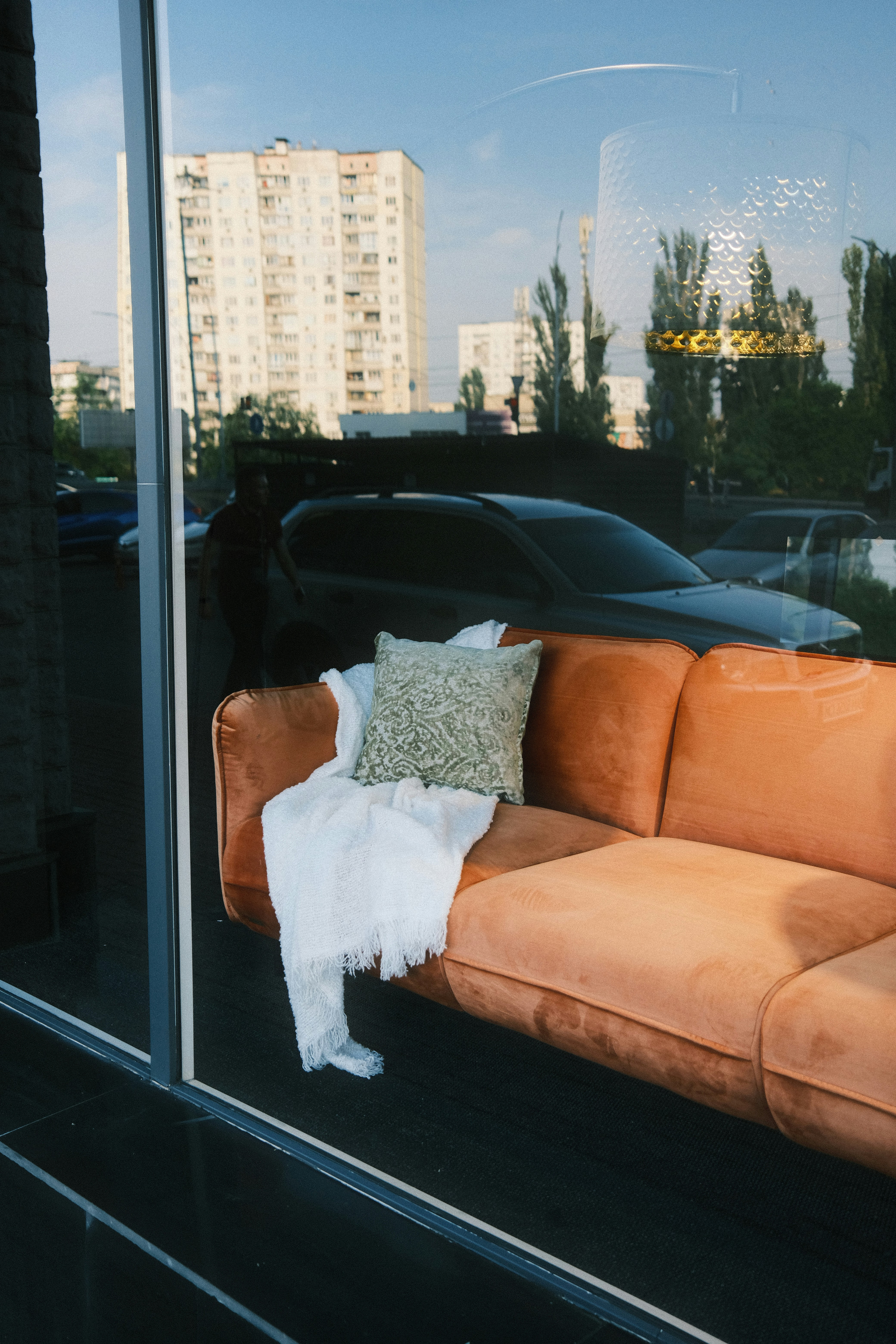 A plush orange couch adorned with a decorative pillow and a white throw, reflecting the urban landscape through a glass window.