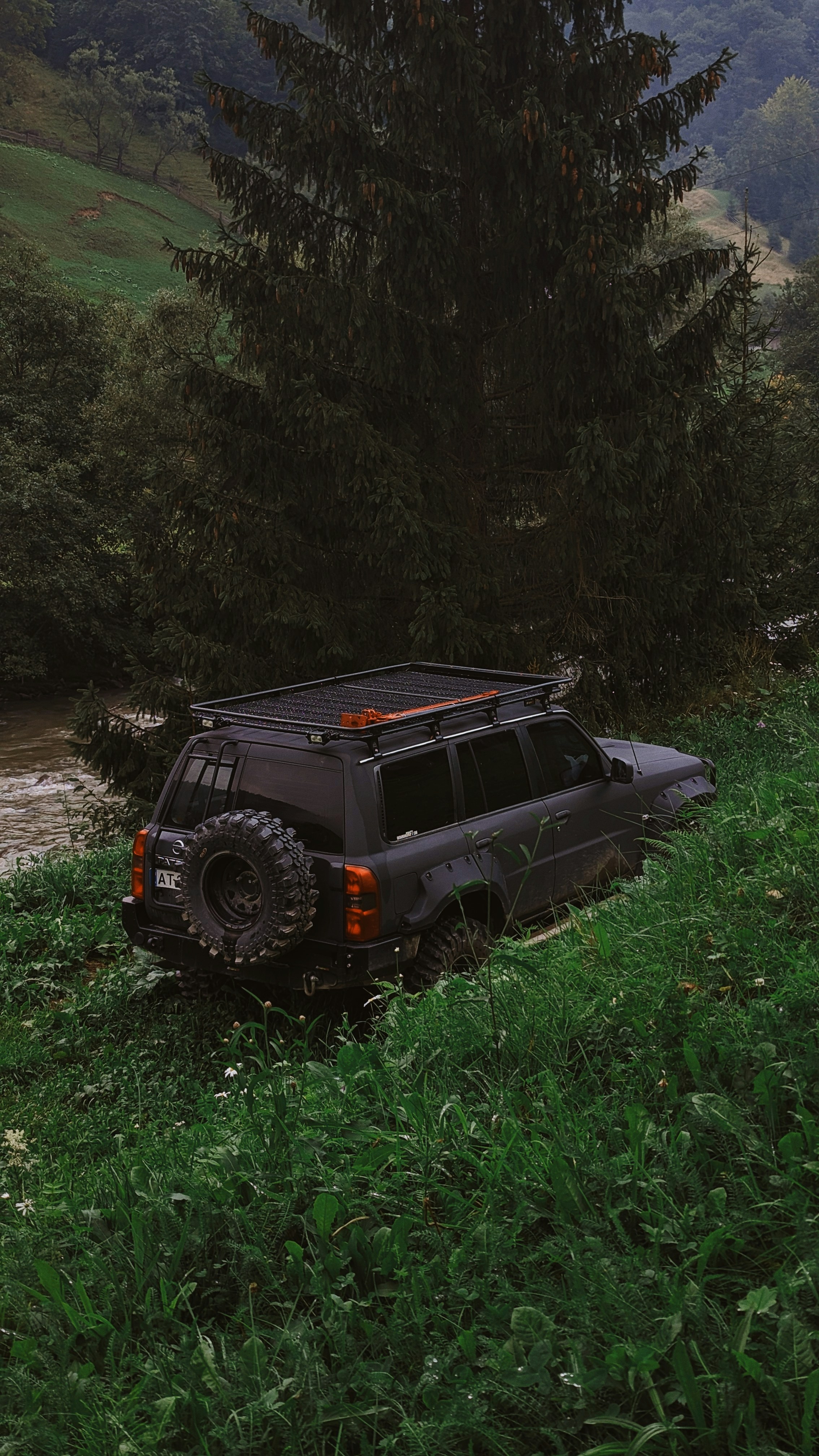 Rugged Quiet: A Dark SUV Nestled by the River Beneath a Conifer