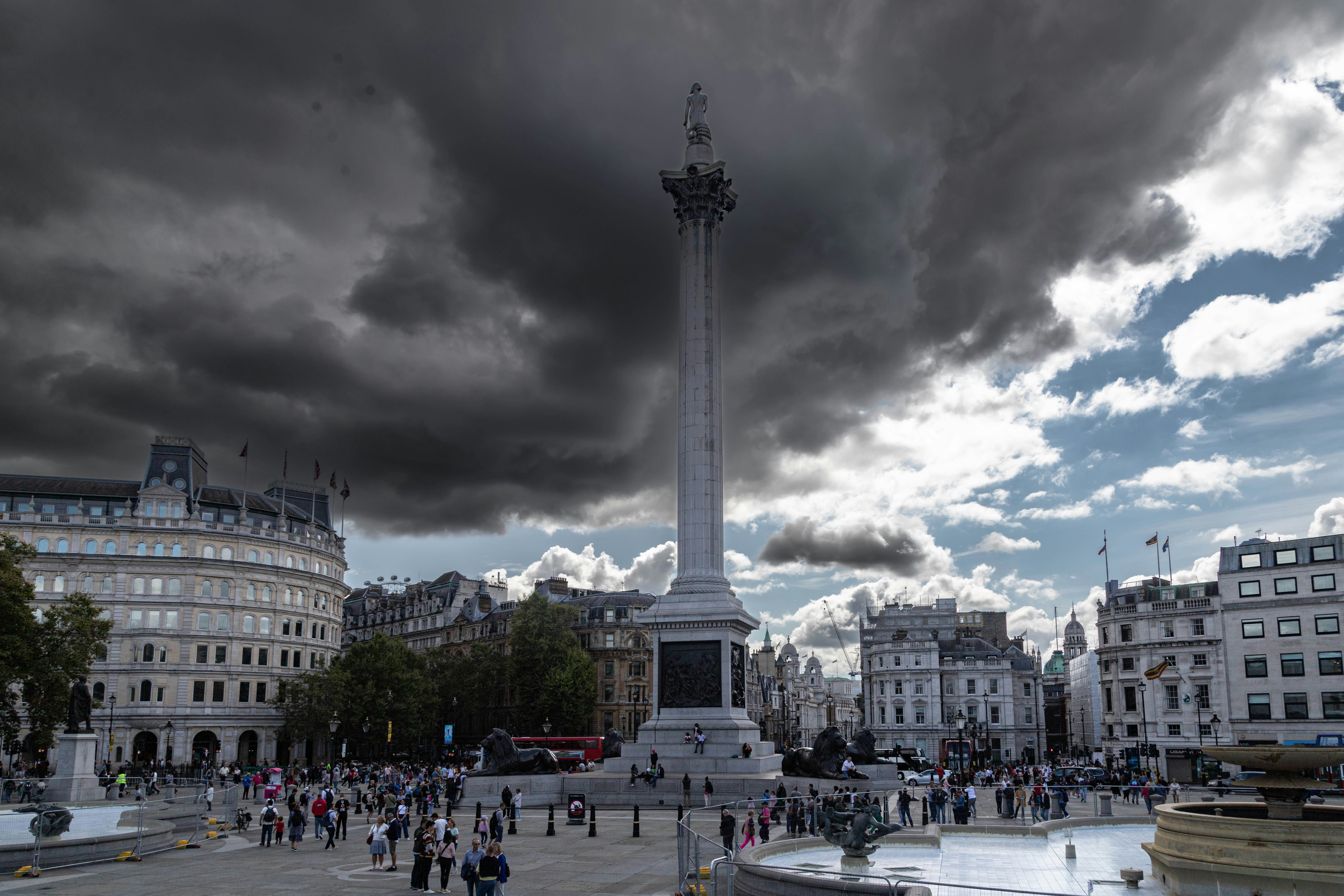 Nelson's Column stands tall in Trafalgar Square, surrounded by crowds and dramatic stormy skies. The scene captures the vibrant atmosphere of a historic landmark.