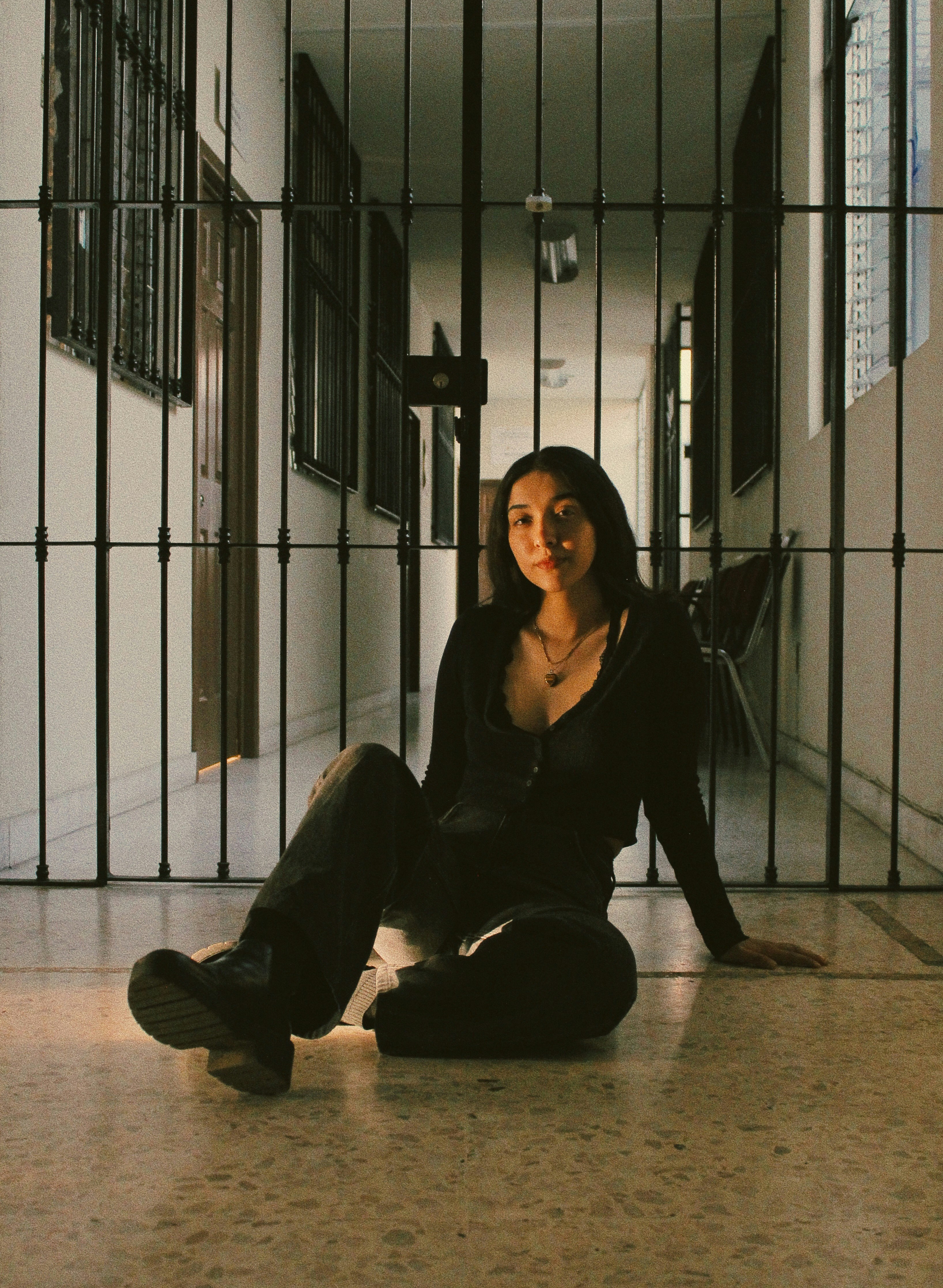 Person seated on a sunlit corridor floor in front of a black metal gate, surrounded by soft shadows.