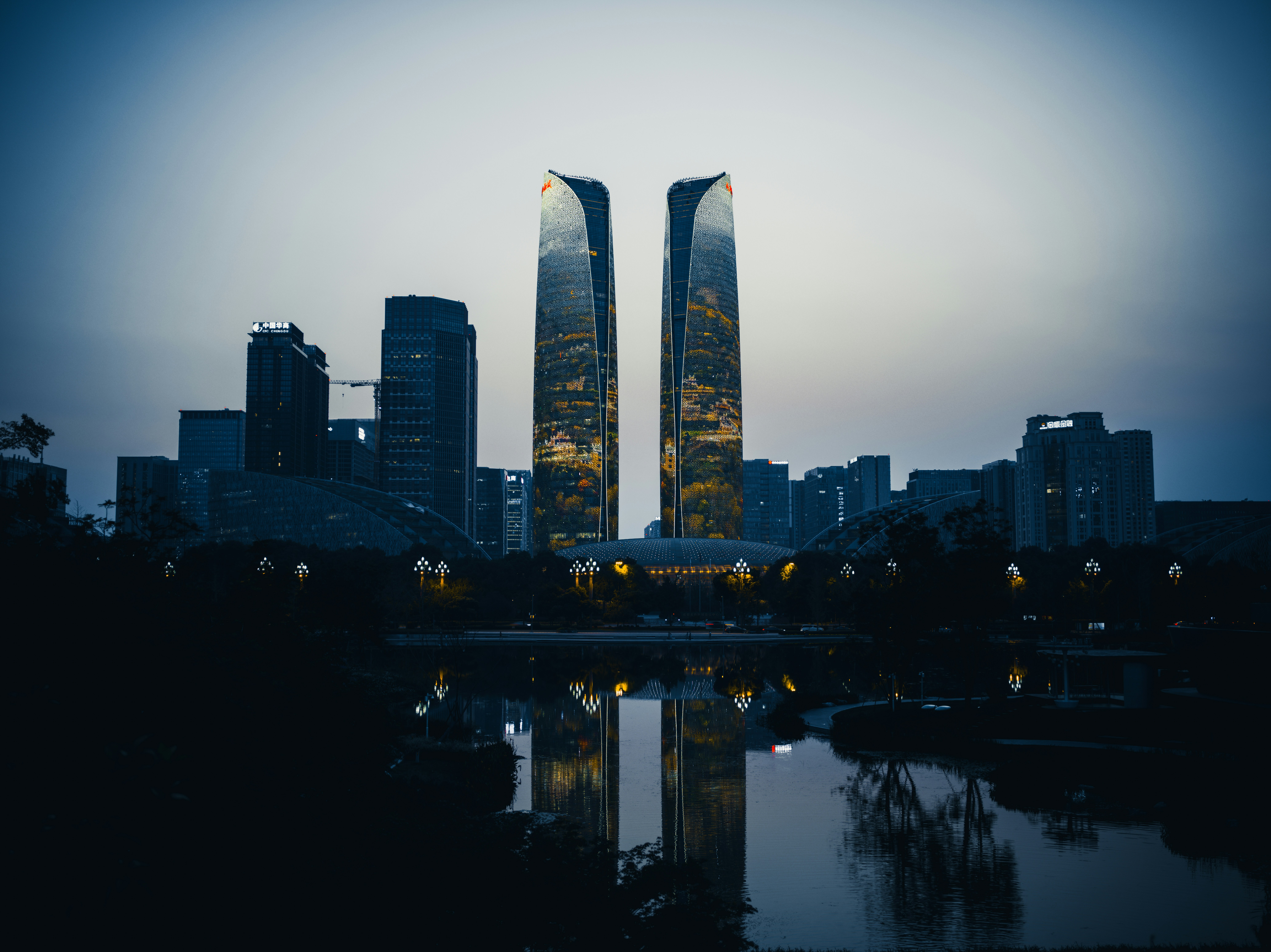 Urban skyline with two illuminated skyscrapers reflecting in a calm body of water at dusk.