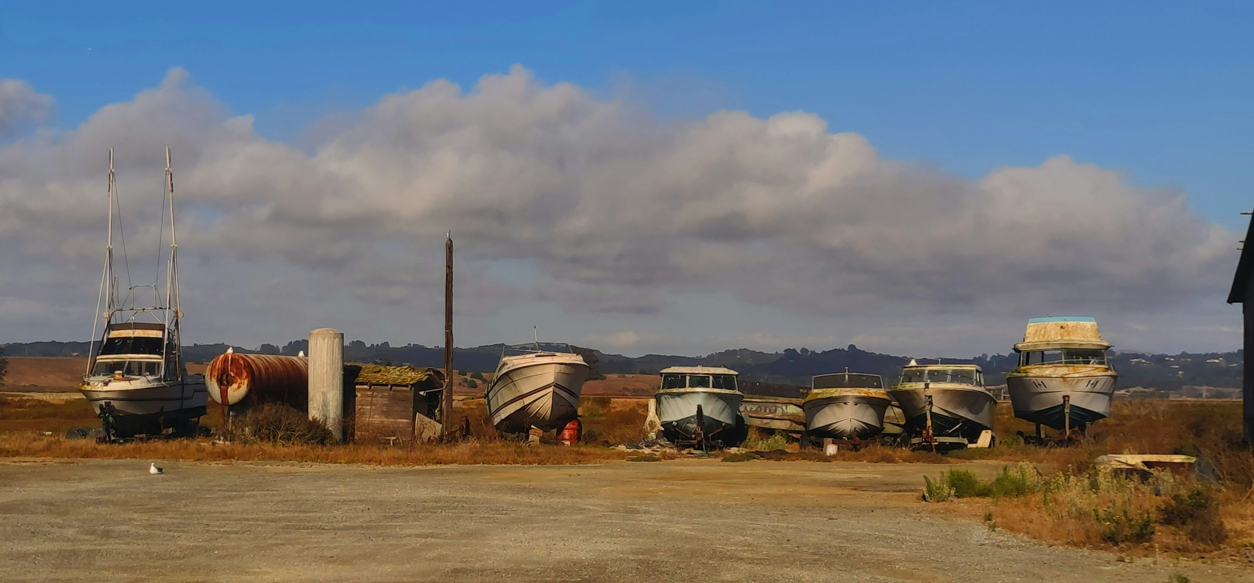 A row of abandoned boats rests on dry land beneath a partly cloudy sky.