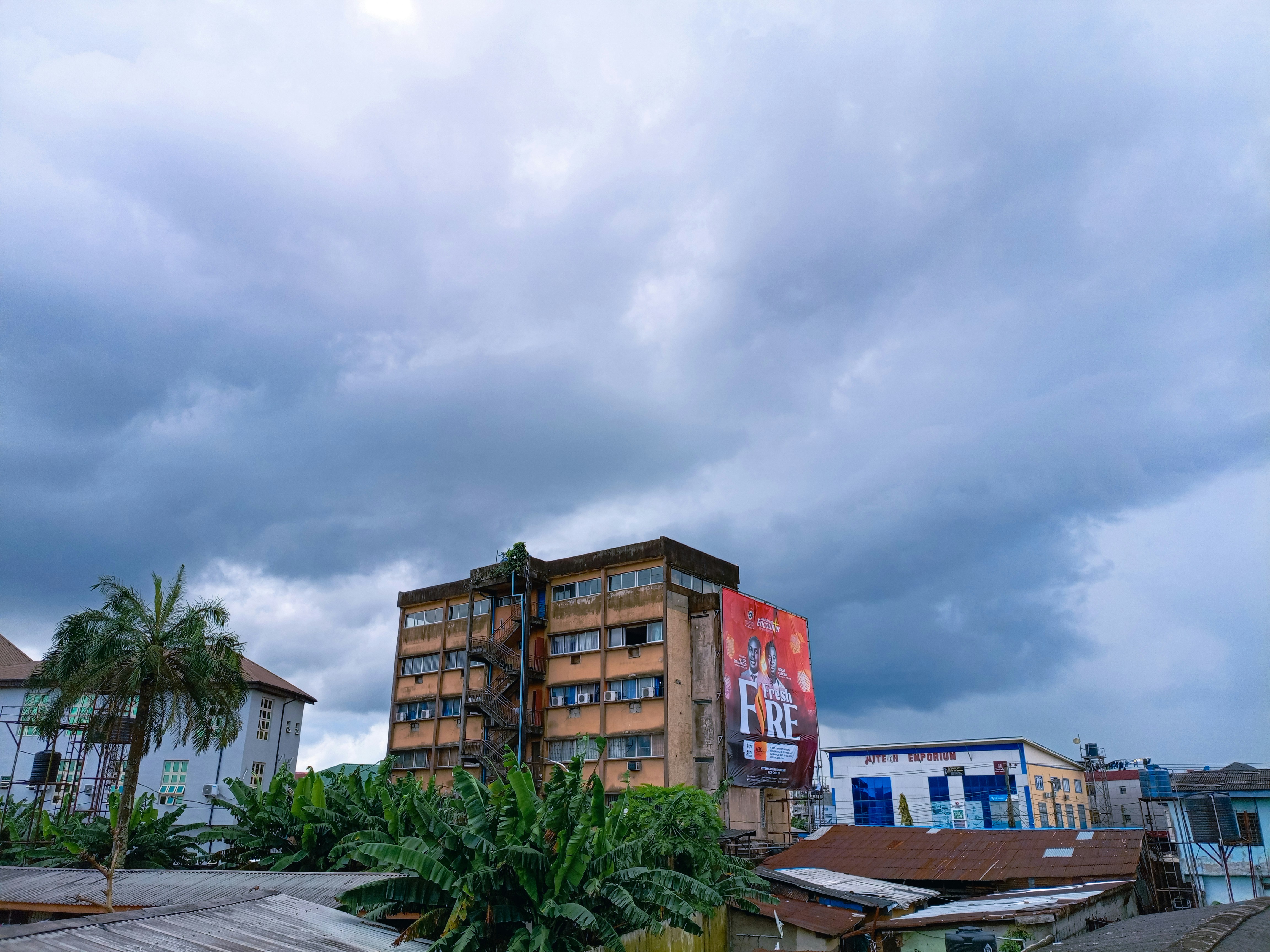 Tall building with vibrant mural beneath dramatic clouds, framed by lush greenery.