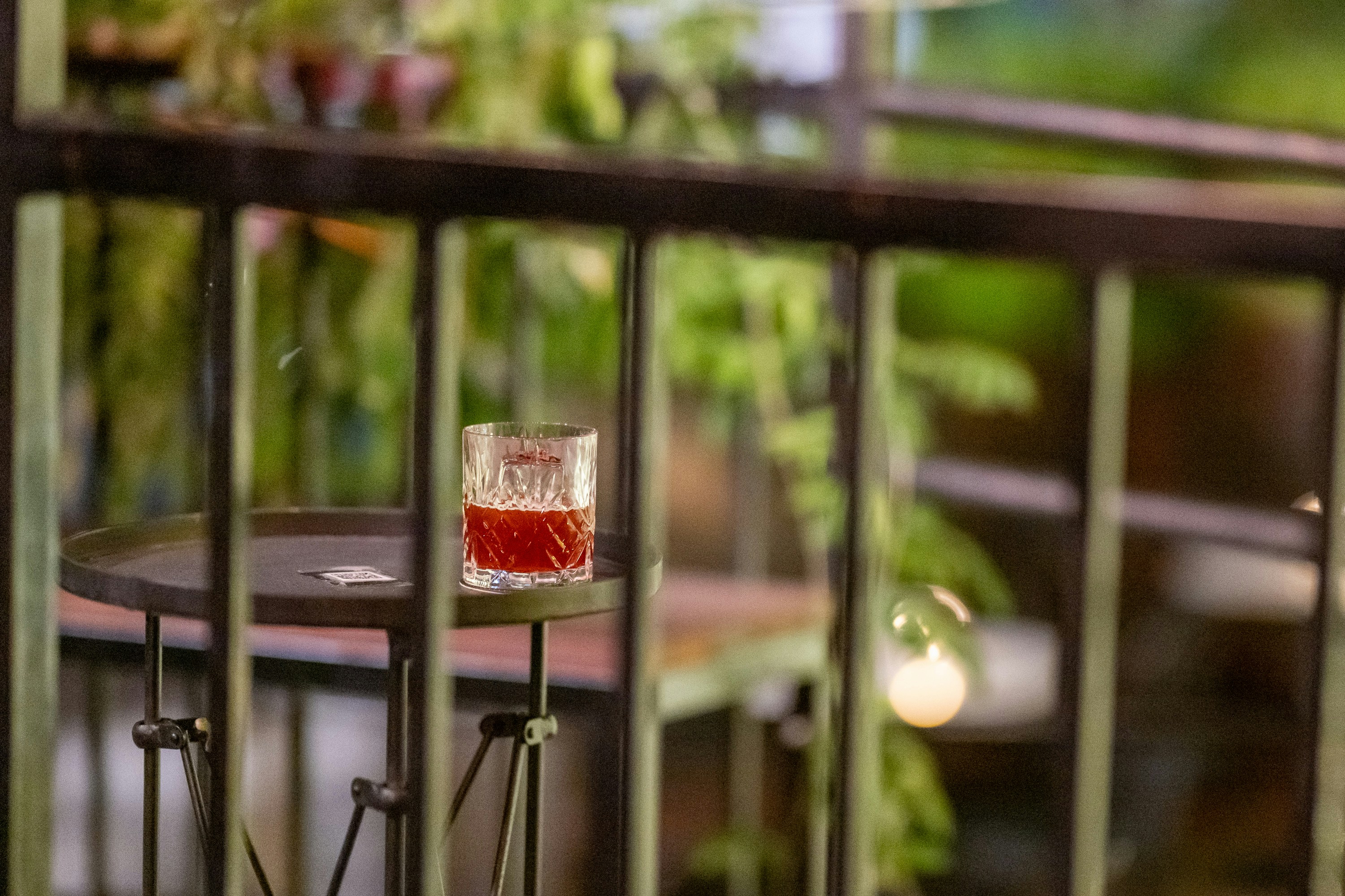 Glass of red cocktail on an outdoor table surrounded by metal railings and lush greenery.