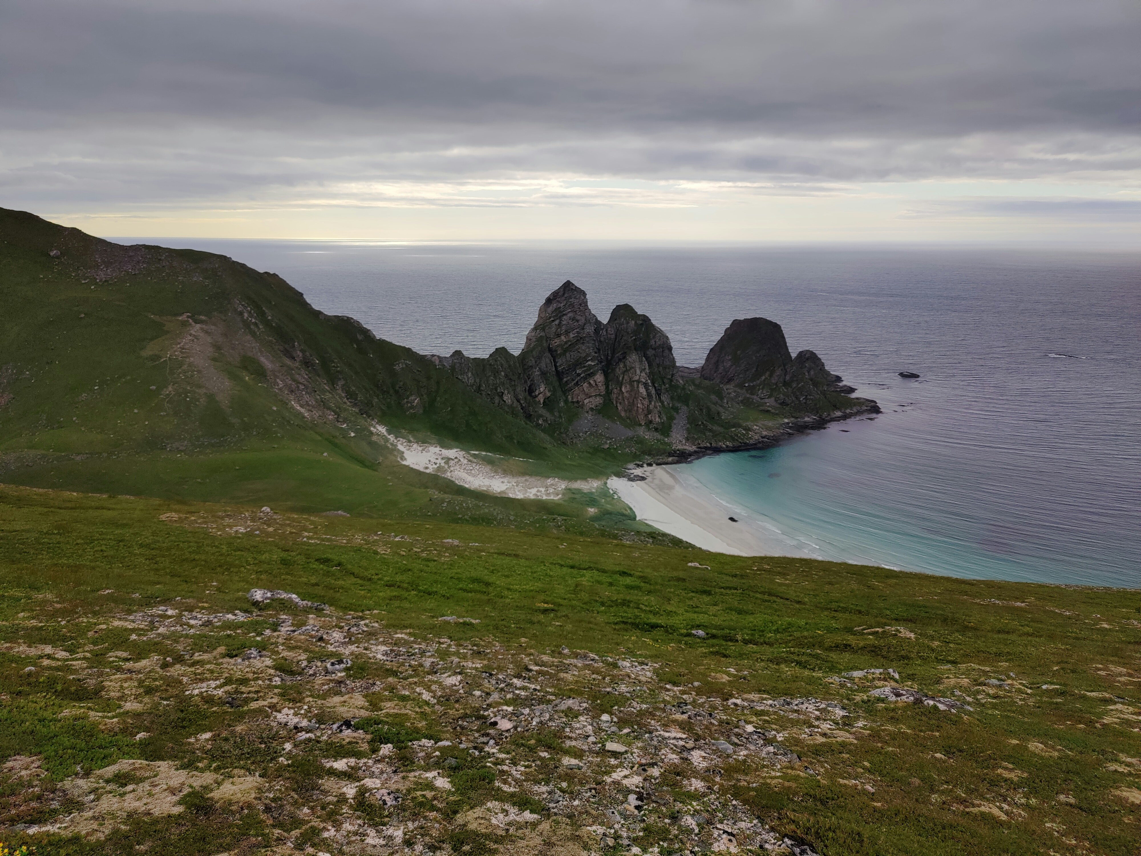Dramatic cliffs rise above a secluded beach under a cloudy sky, with a calm sea stretching to the horizon.