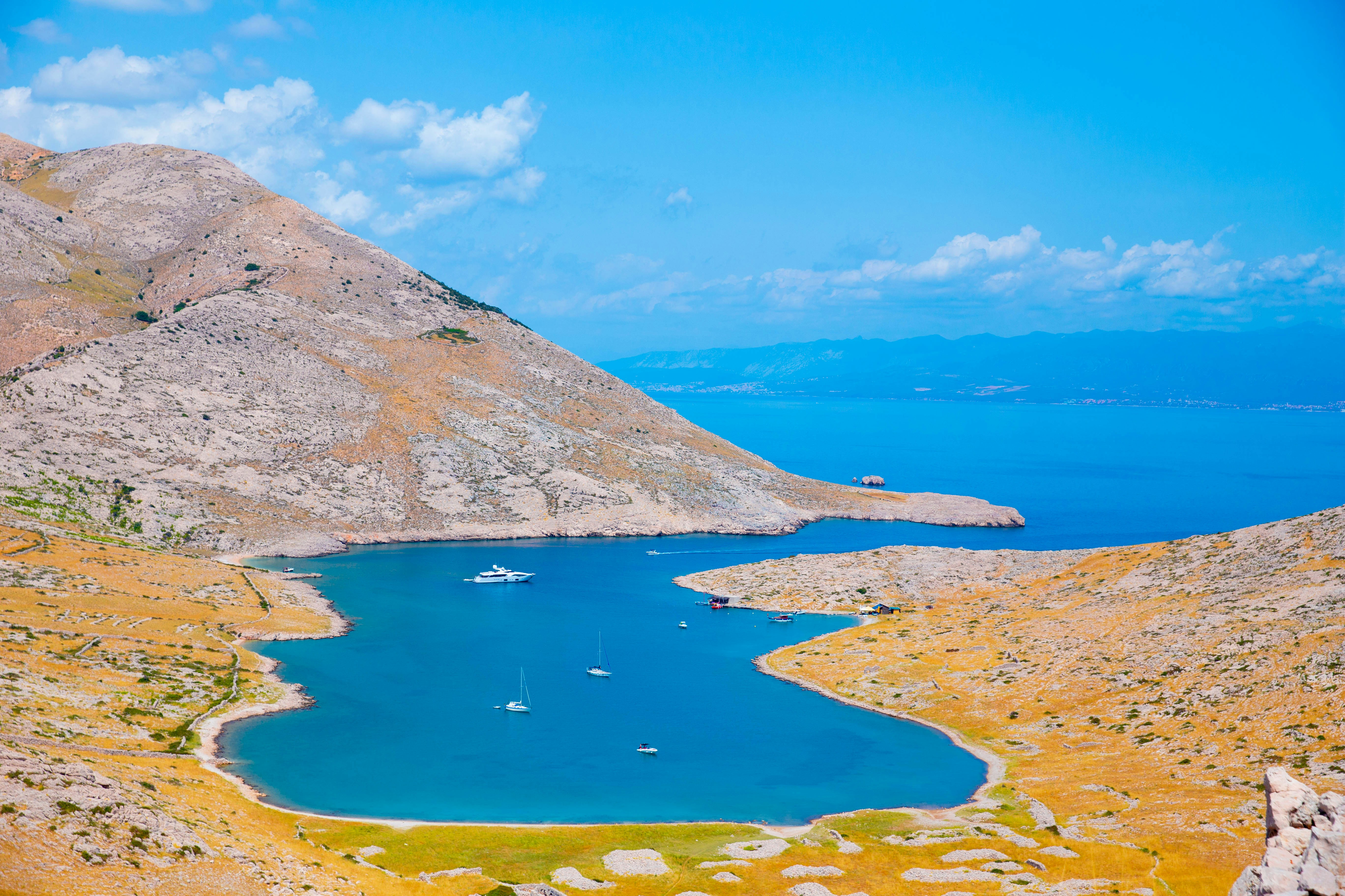 A tranquil bay surrounded by dry, rugged hills and dotted with sailboats under a bright blue sky.