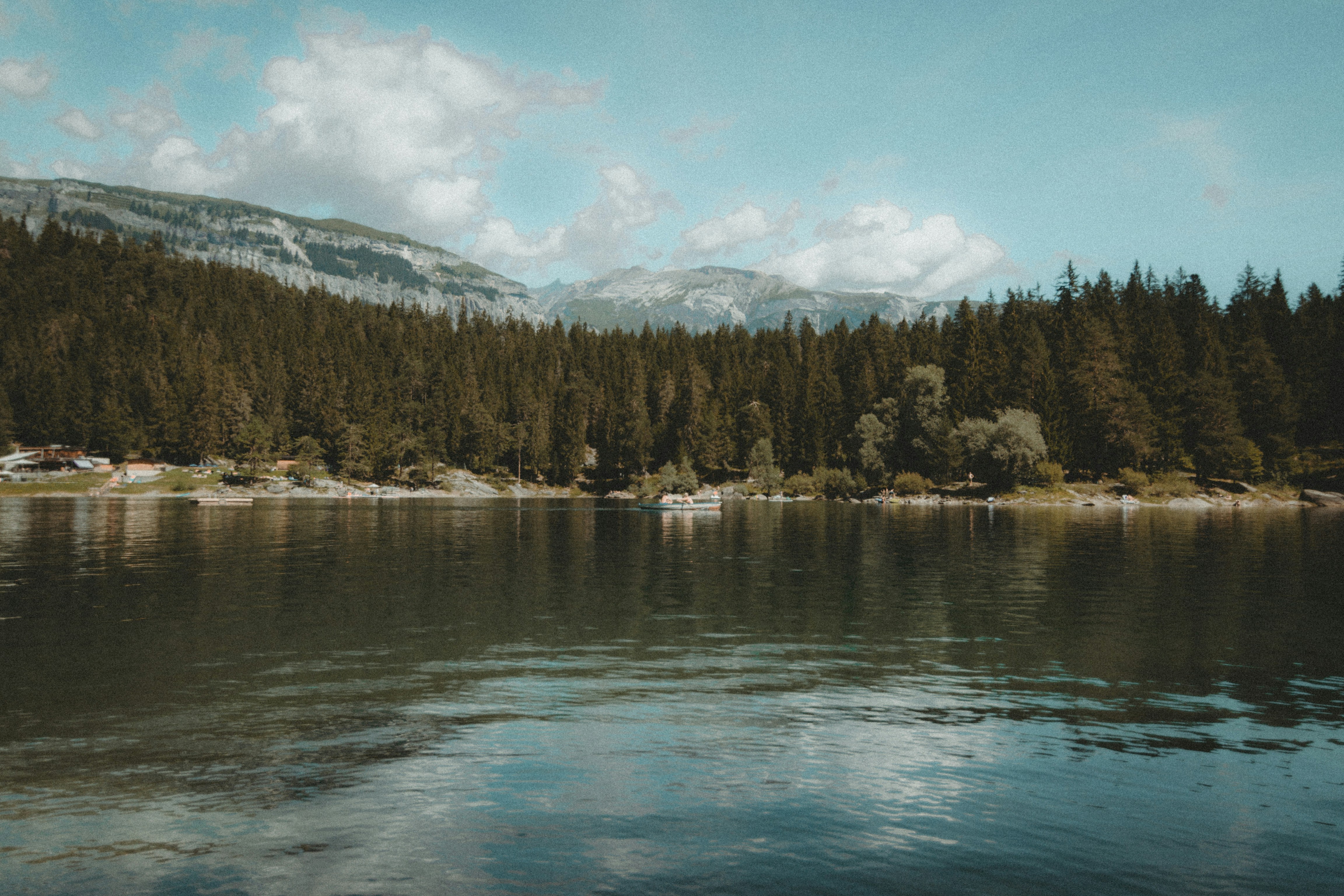 Tranquil lake reflecting a dense forest and distant mountains under a partly cloudy sky.