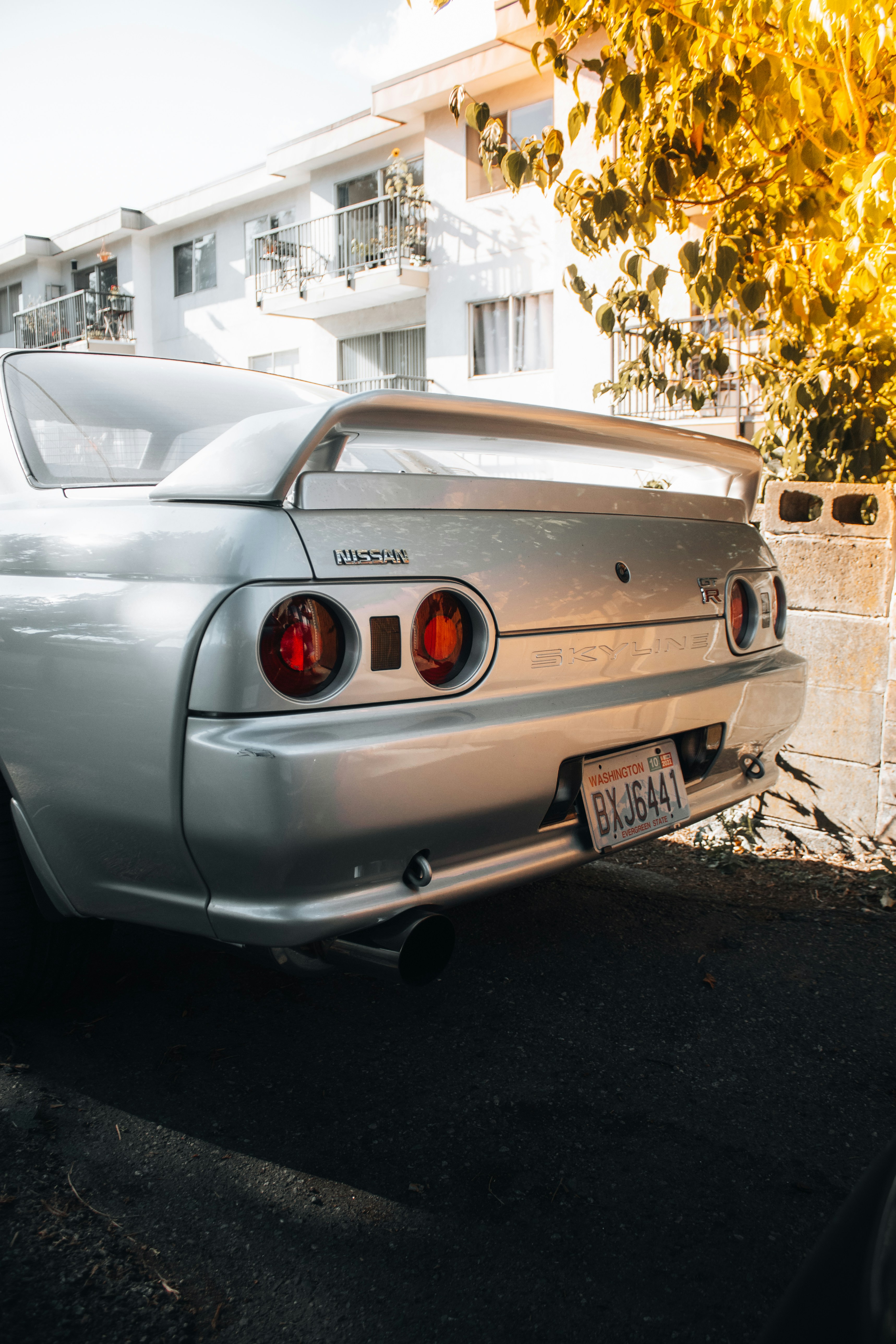 Silver car with a spoiler parked near an apartment building under a tree with golden leaves.