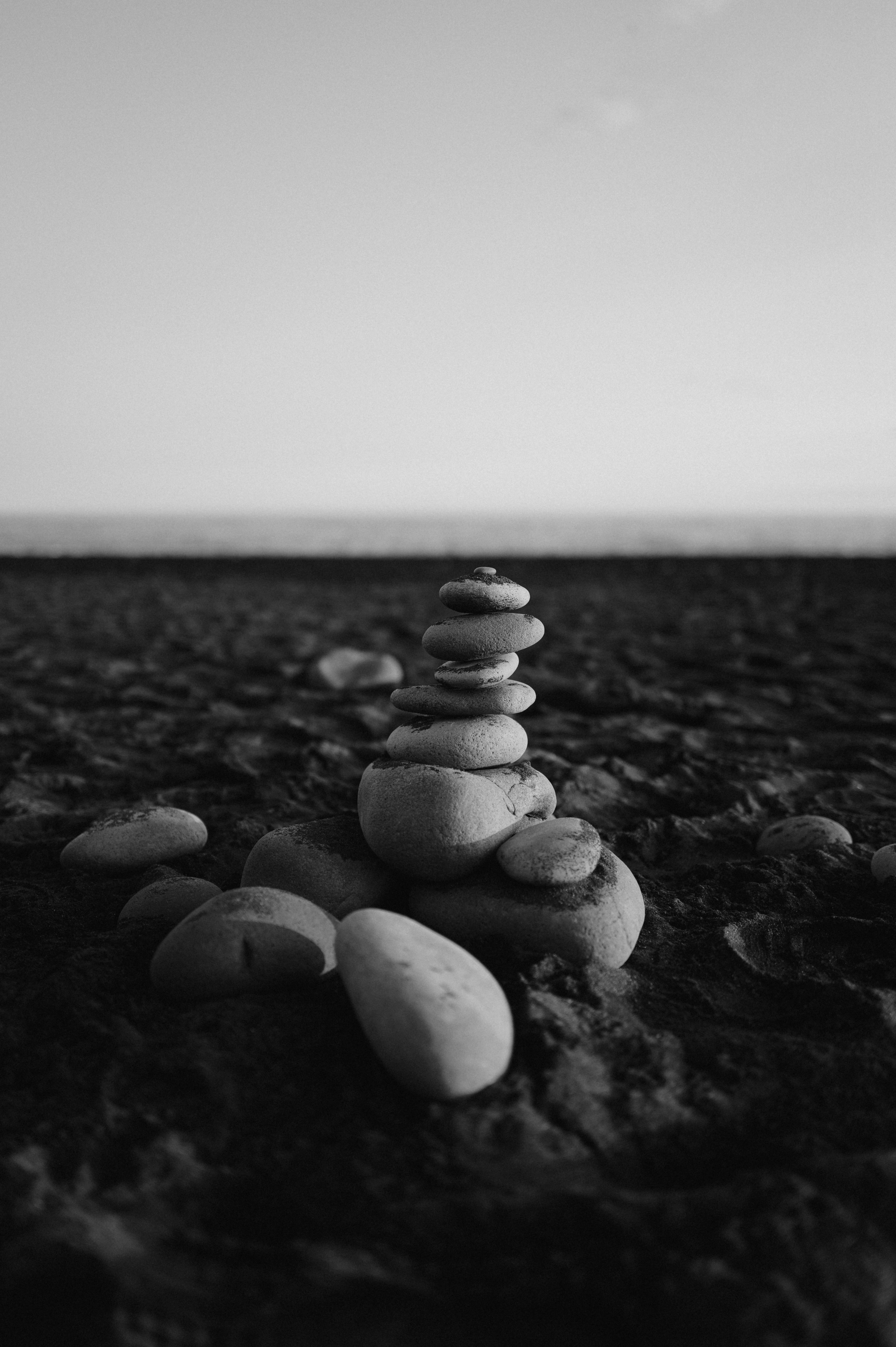Monochrome photograph of a compact stack of smooth stones on a textured beach, with a distant horizon. The composition emphasizes balance and stillness.