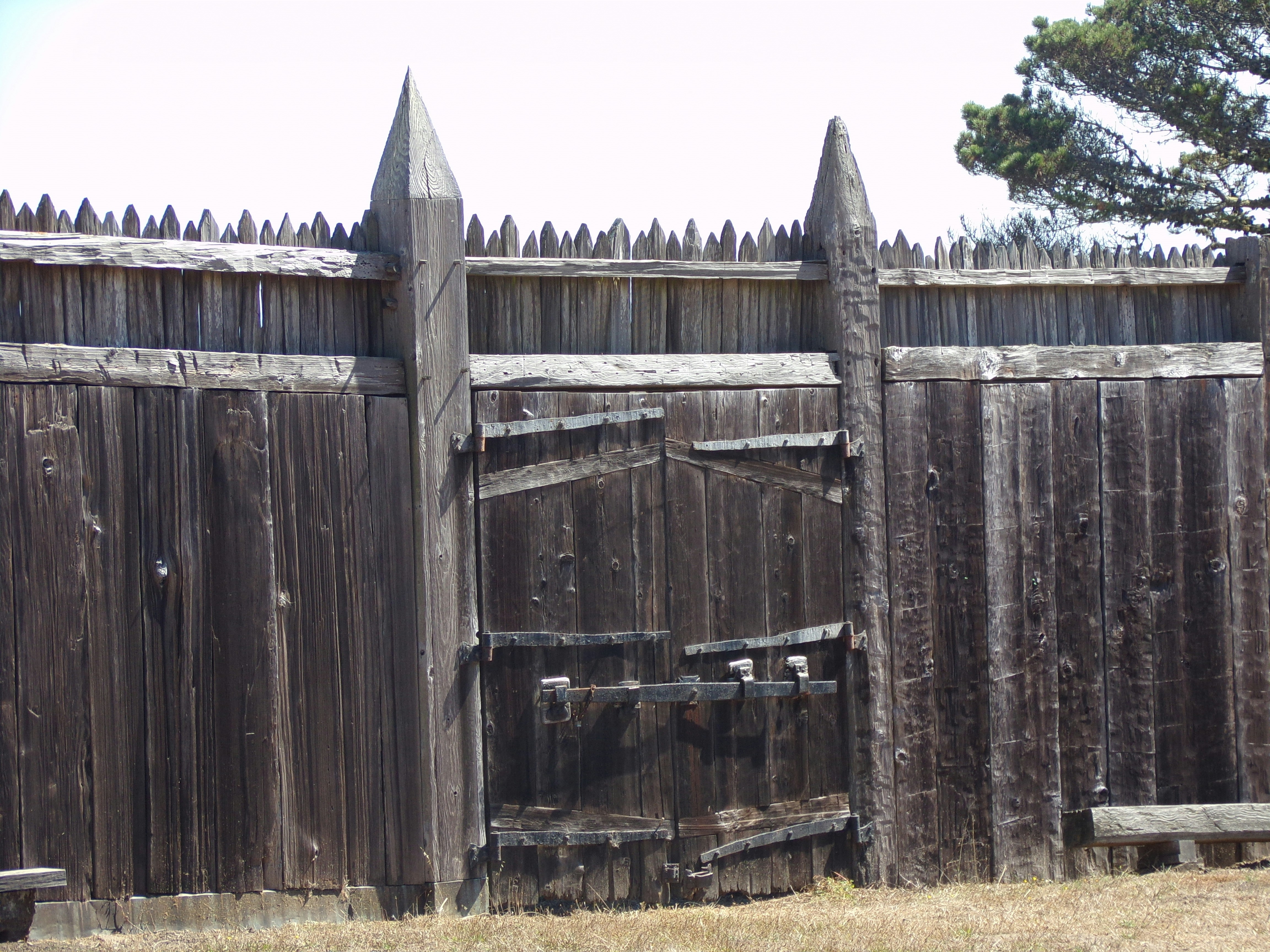 Photograph of a weathered wooden fence with a central gate, bathed in daylight. The shot emphasizes the rustic barrier and its worn texture.